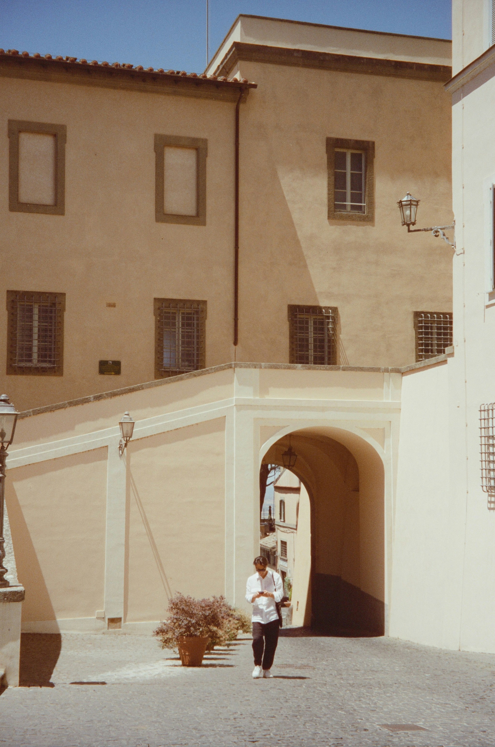 A person walks beneath a sunlit archway in a courtyard of warm-toned buildings; the scene emphasizes the architectural scale and a single figure for contrast. This is a photograph.