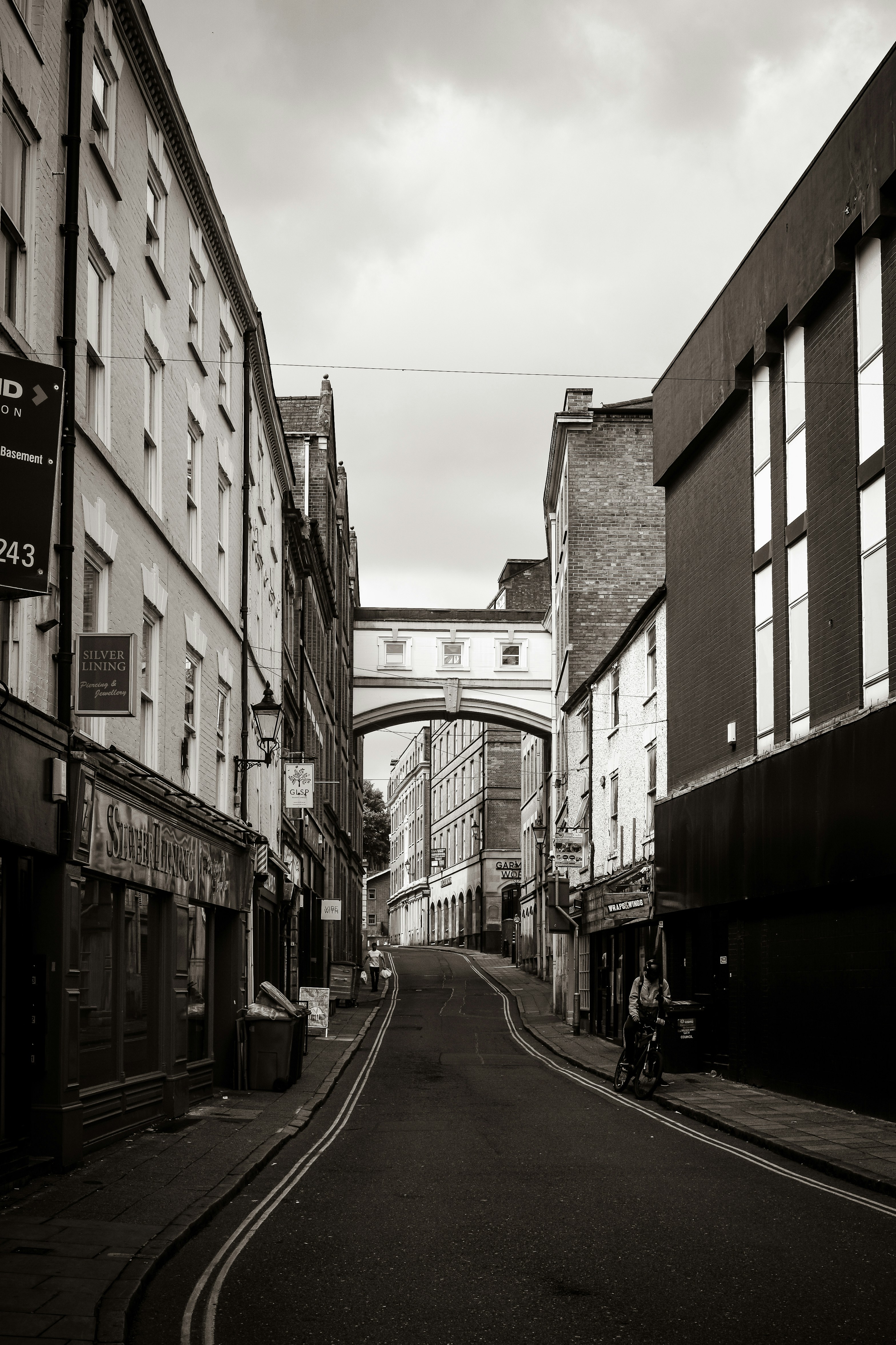 Monochrome street scene of a narrow city lane climbing toward an arched bridge linking brick façades, with a cyclist waiting by the curb.