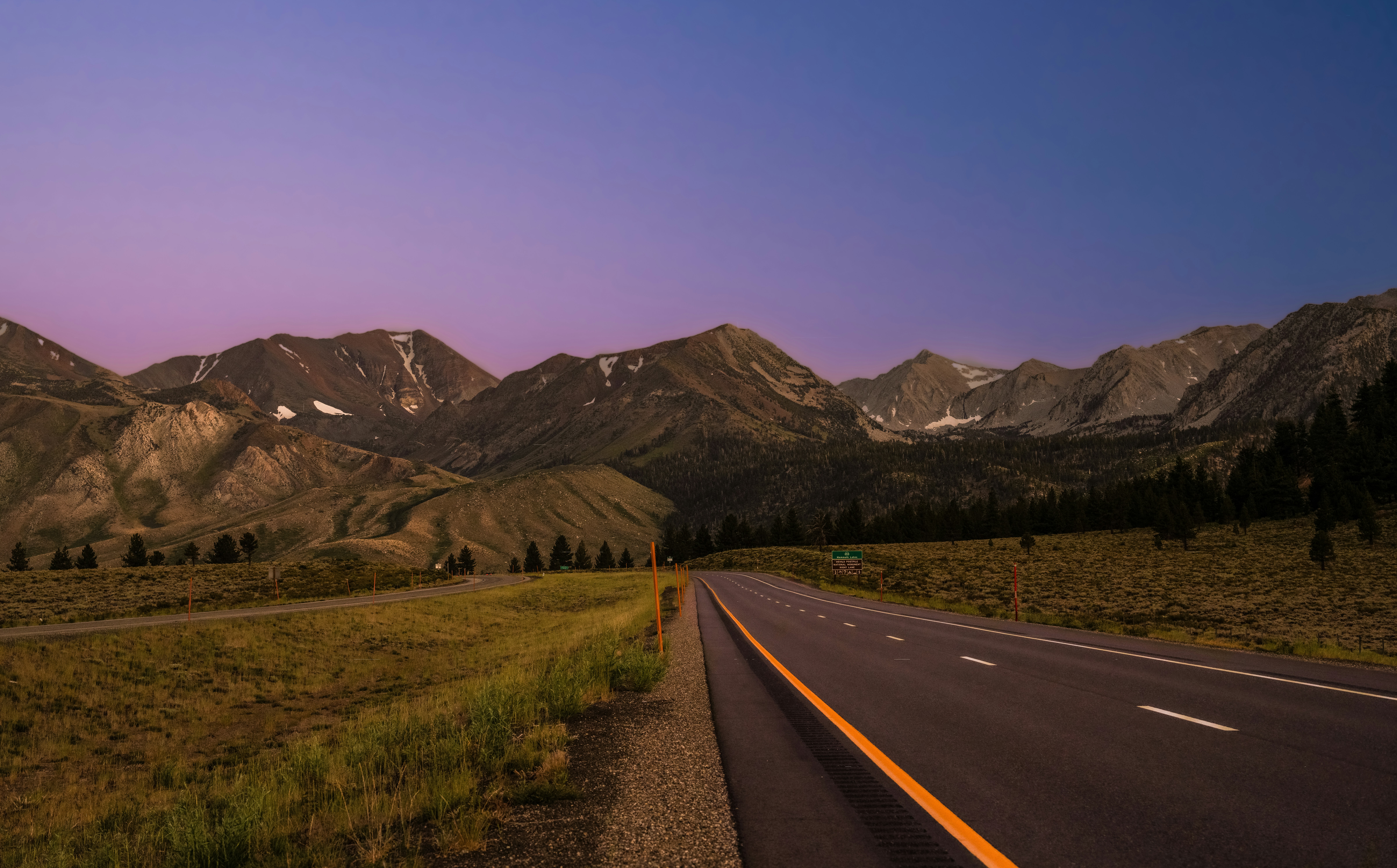 Winding highway bordered by lush grasslands, leading toward majestic mountain ranges under a twilight sky.