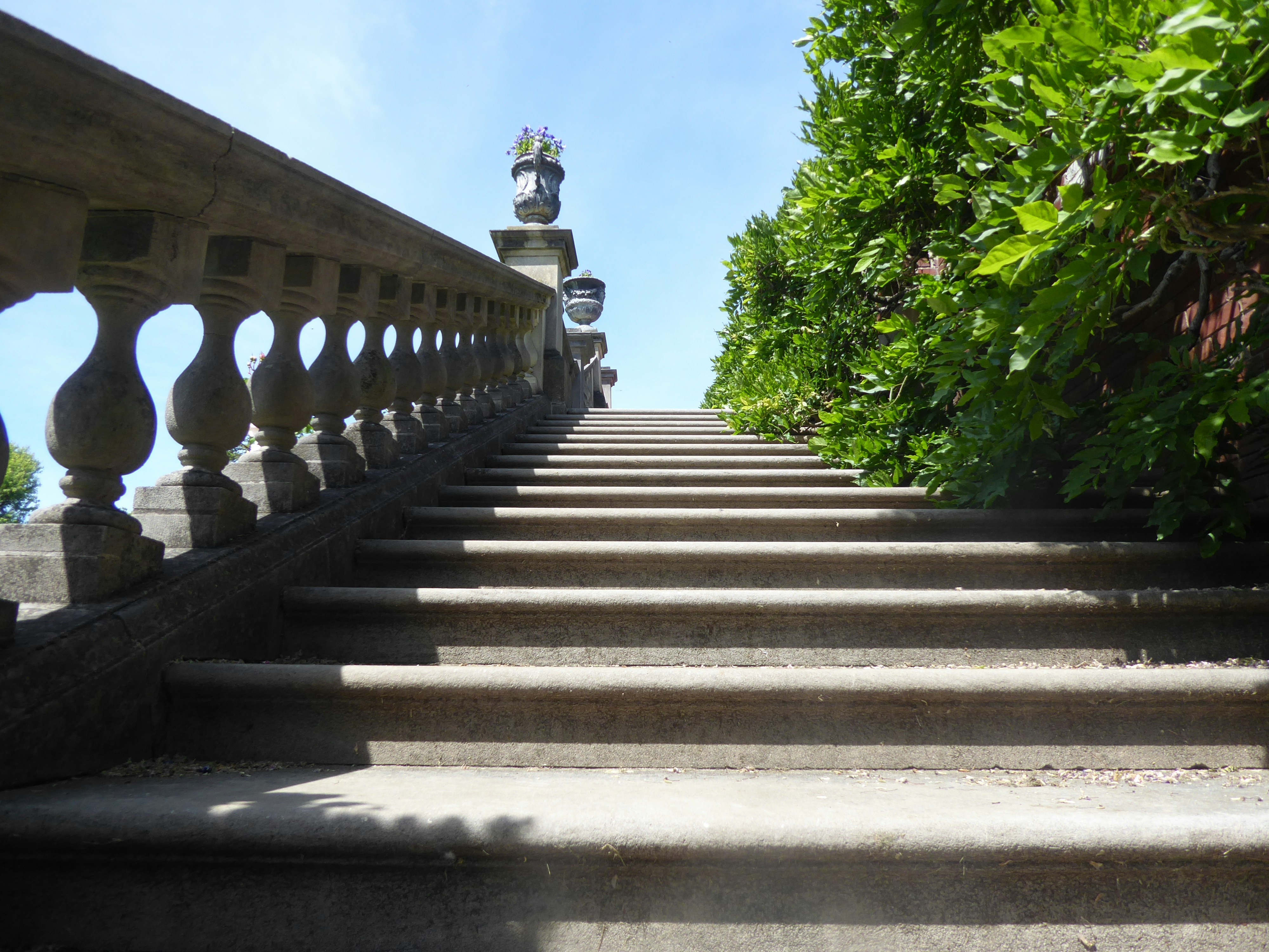 Stone staircase with ornate balustrade on the left and lush hedge on the right leads upward toward a bright blue sky.