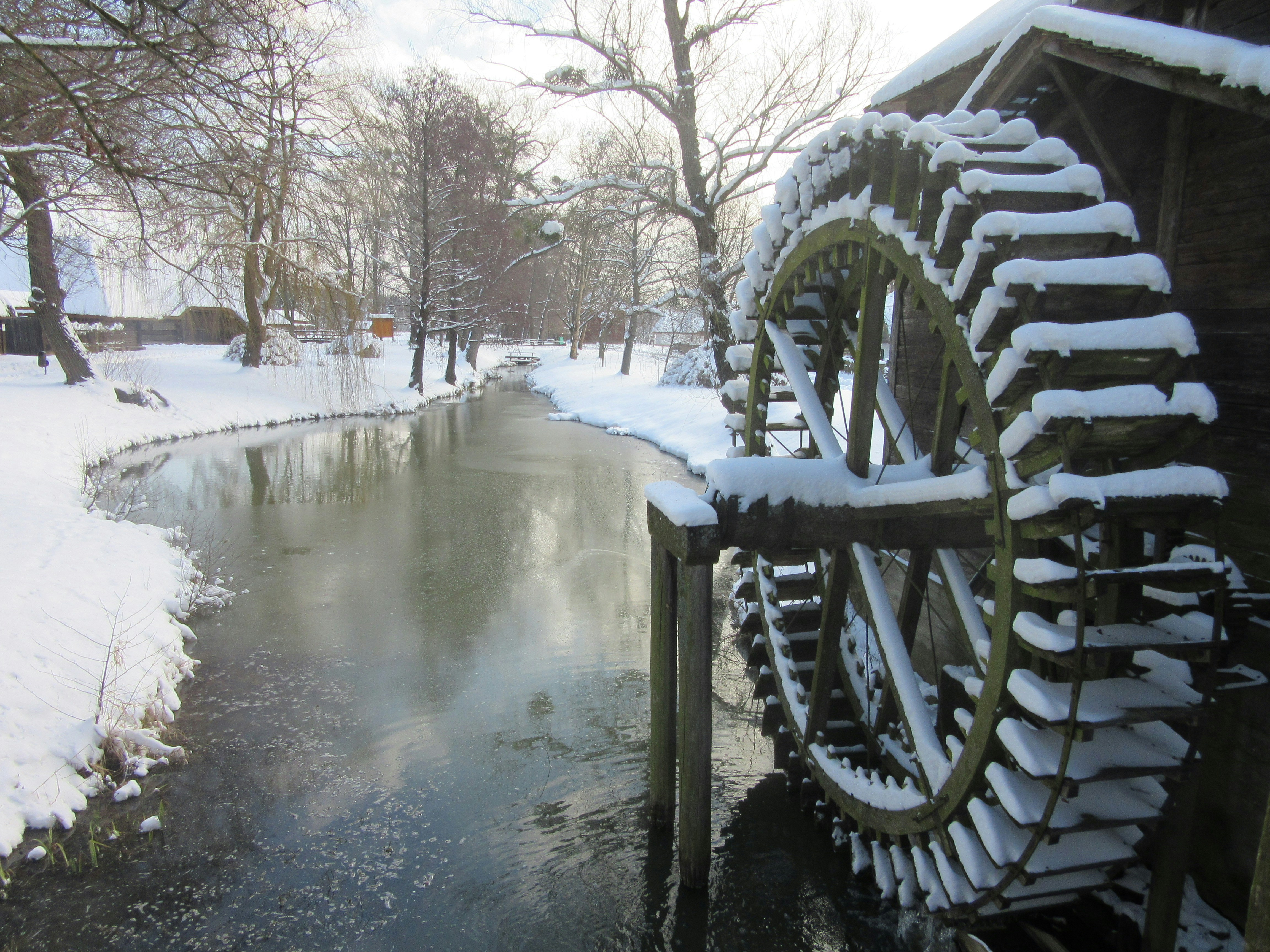 A snow-covered waterwheel stands beside a tranquil stream, framed by frosted trees in a serene winter landscape.