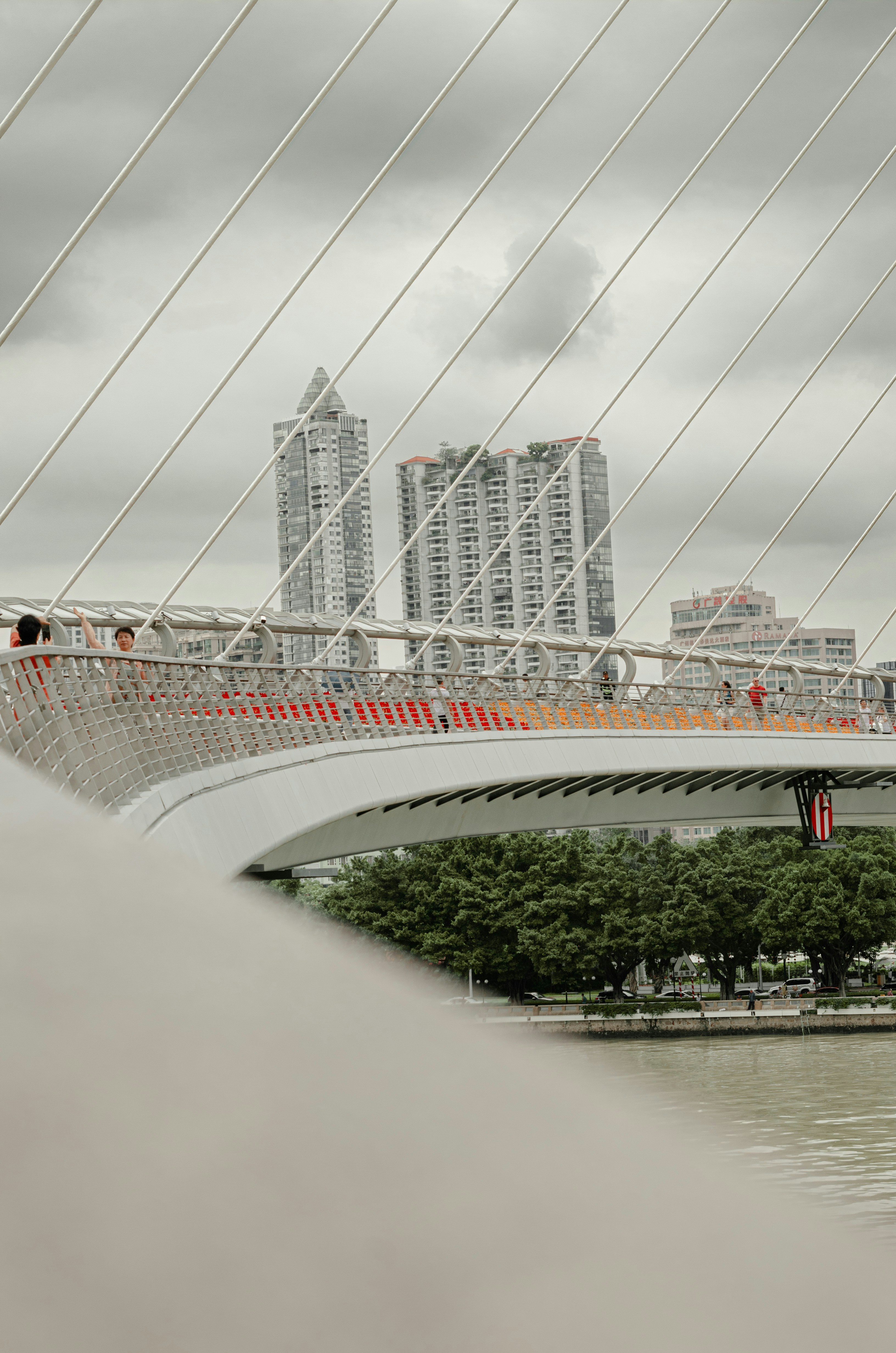 Pedestrian bridge with diagonal steel cables arches over a river, city high-rises visible in the background and trees along the bank.