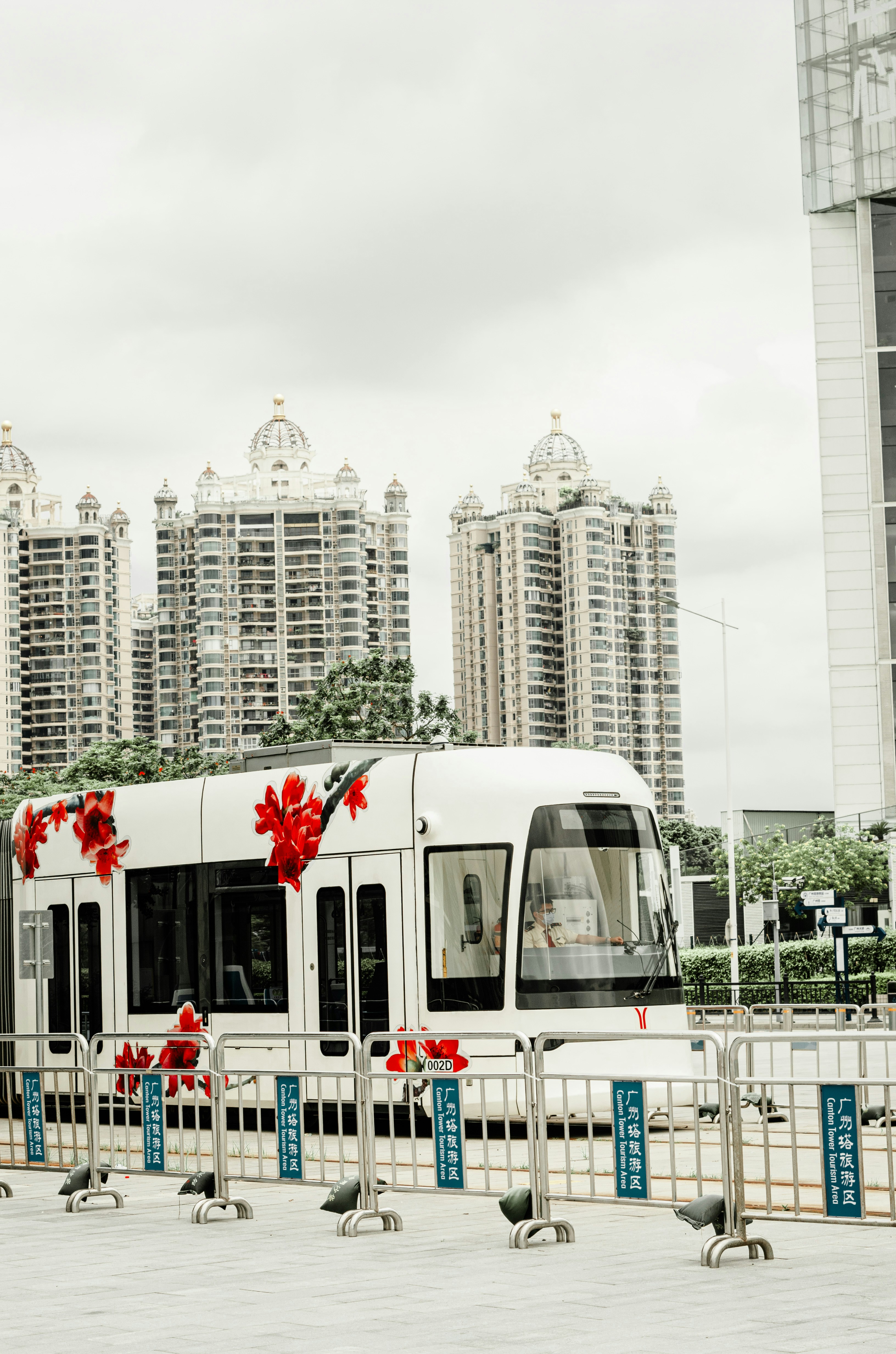 A white tram adorned with red flower graphics sits in a fenced urban plaza, with a skyline of tall ornate towers behind it.