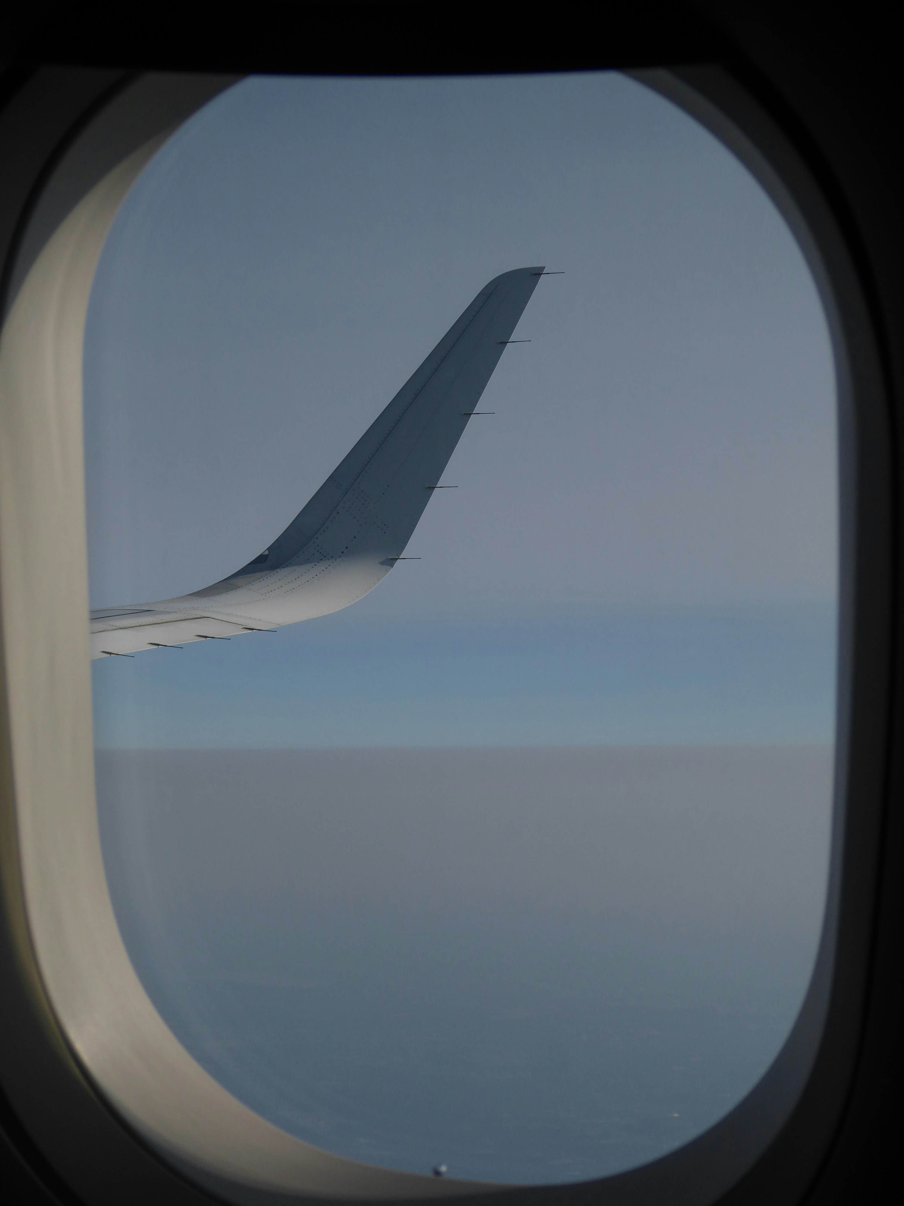 Photograph of a winglet seen through an airplane window, with a pale blue sky and distant ocean below.