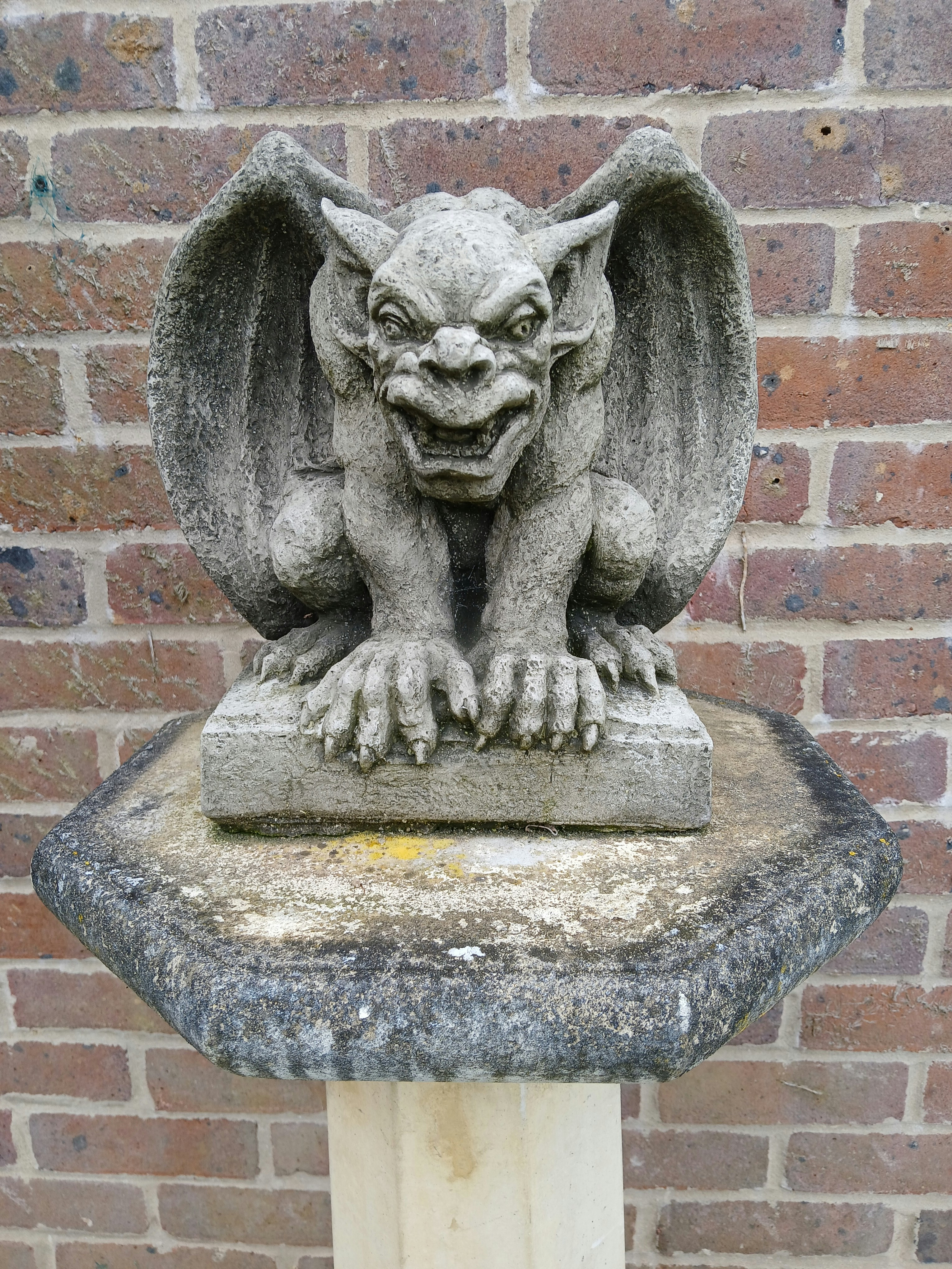 Weathered stone gargoyle statue with wings crouches on a pedestal against a brick wall, its claws gripping the base with a menacing grin.