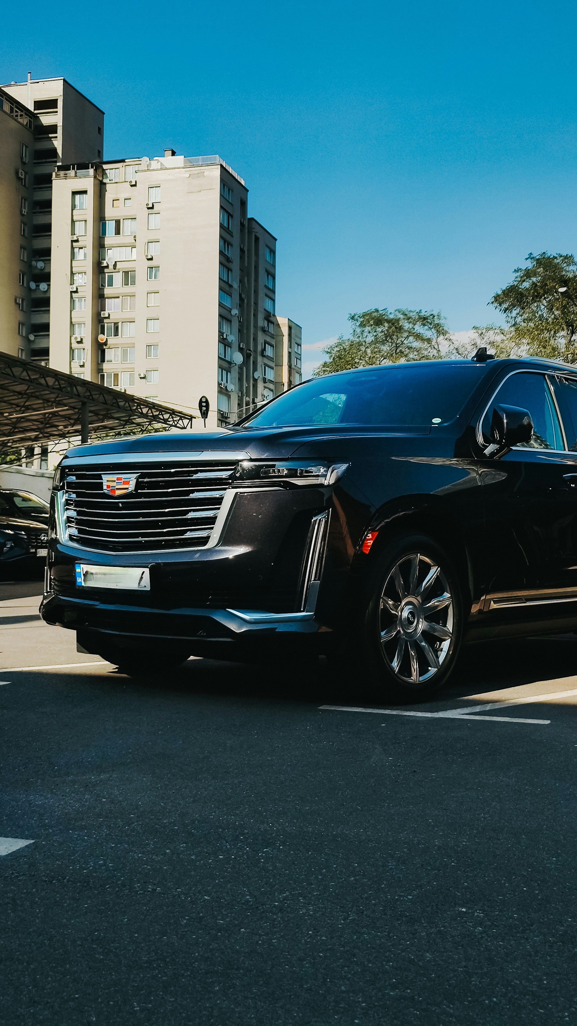 A black Cadillac SUV sits prominently in a sunlit urban lot, its chrome grille catching the light with a glossy finish. Tall apartment buildings and a clear blue sky form the backdrop, emphasizing the vehicle's presence in the city.