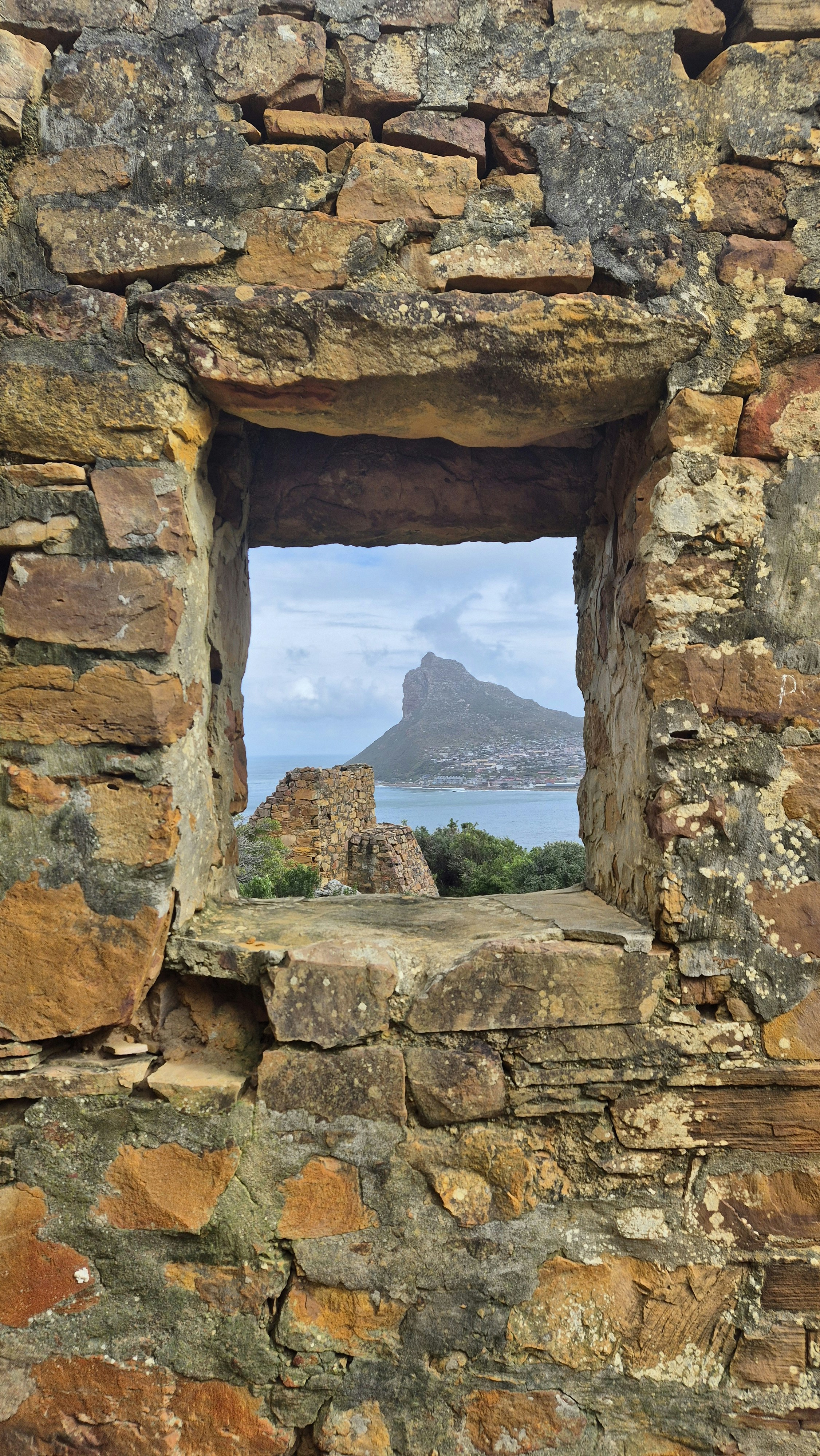 Rugged stone wall with a rectangular window frames a distant island and sea. The composition emphasizes the view through an ancient ruin.