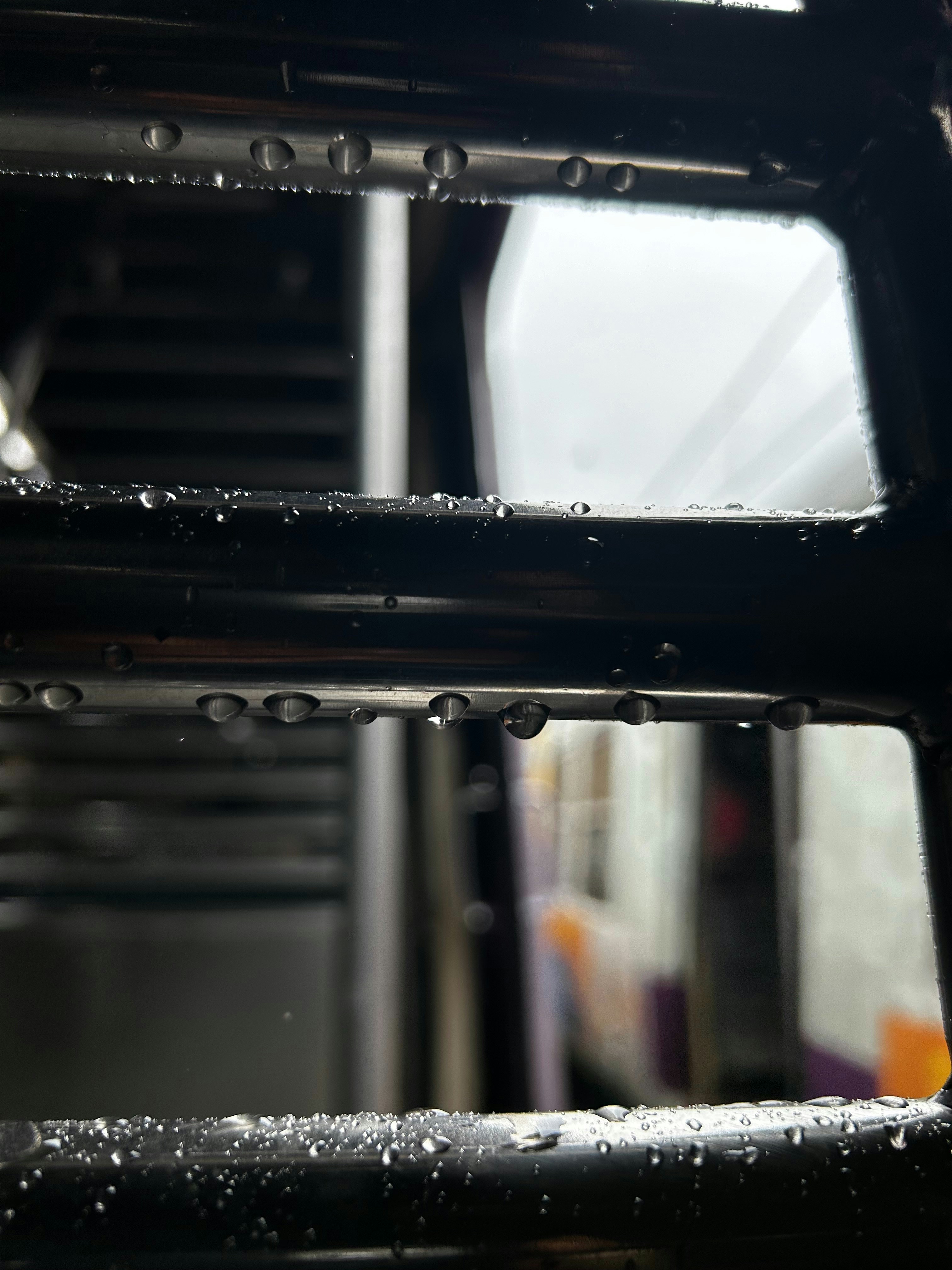 Close-up photograph of a rain-speckled black steel grate with droplets bead along the horizontal slats; background softly blurred.