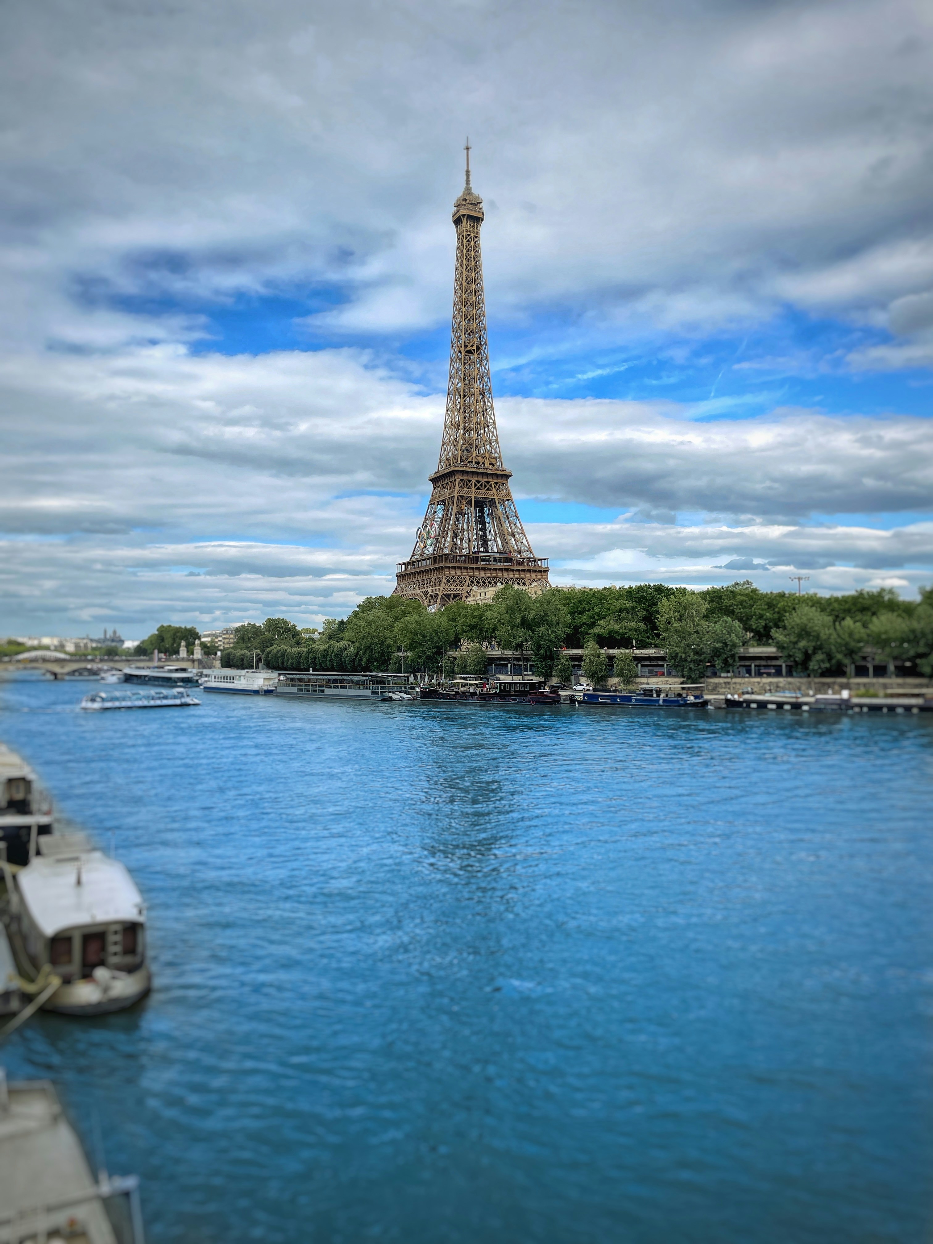 Seine riverfront with the Eiffel Tower rising above green banks and boats docked along the quay under a partly cloudy sky.