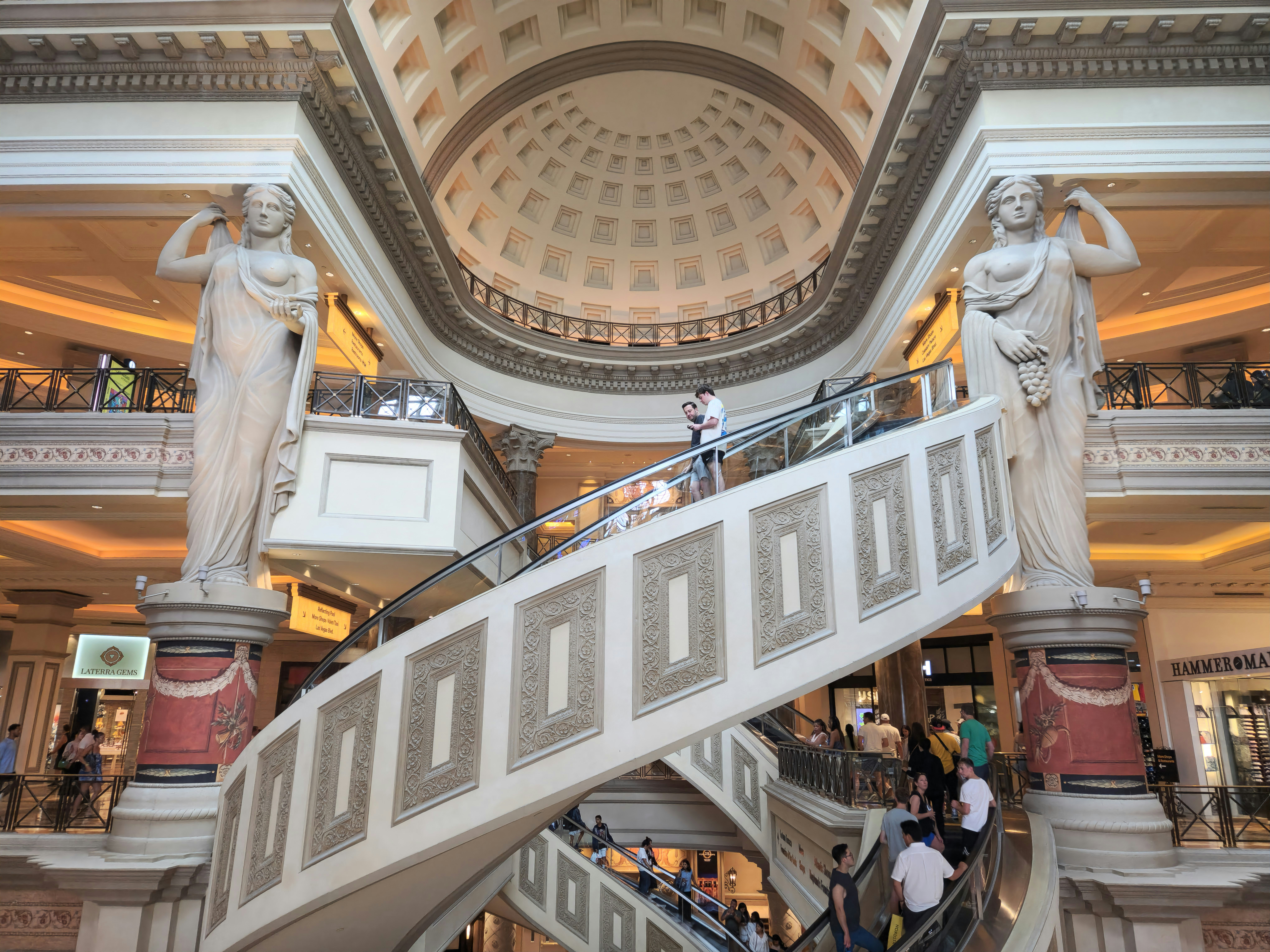 Spiral staircase framed by classical sculptures in a lavish atrium, showcasing intricate architectural details and lively activity below.