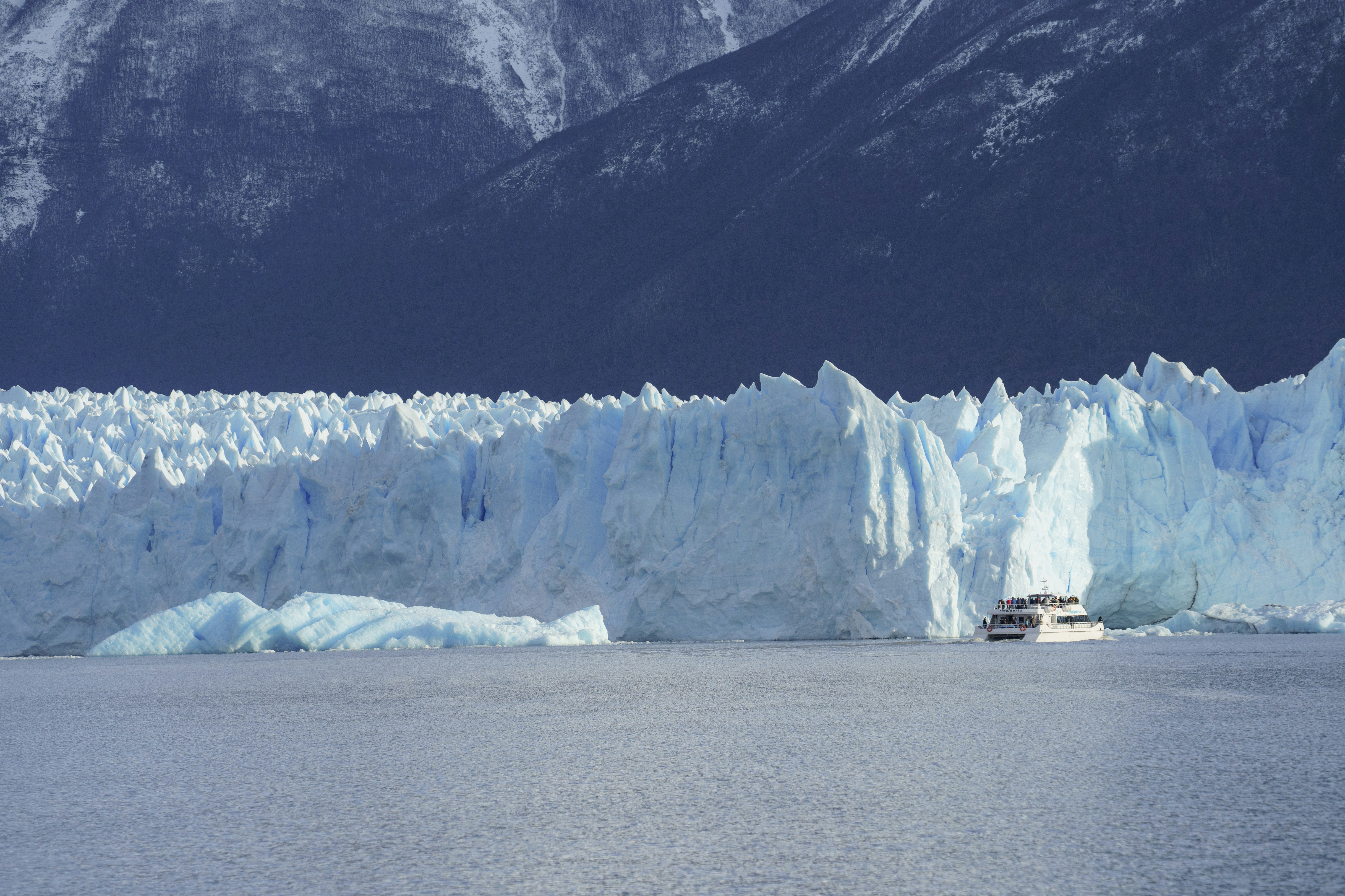 Vast glacier with rugged icy walls set against towering dark mountains and calm waters.