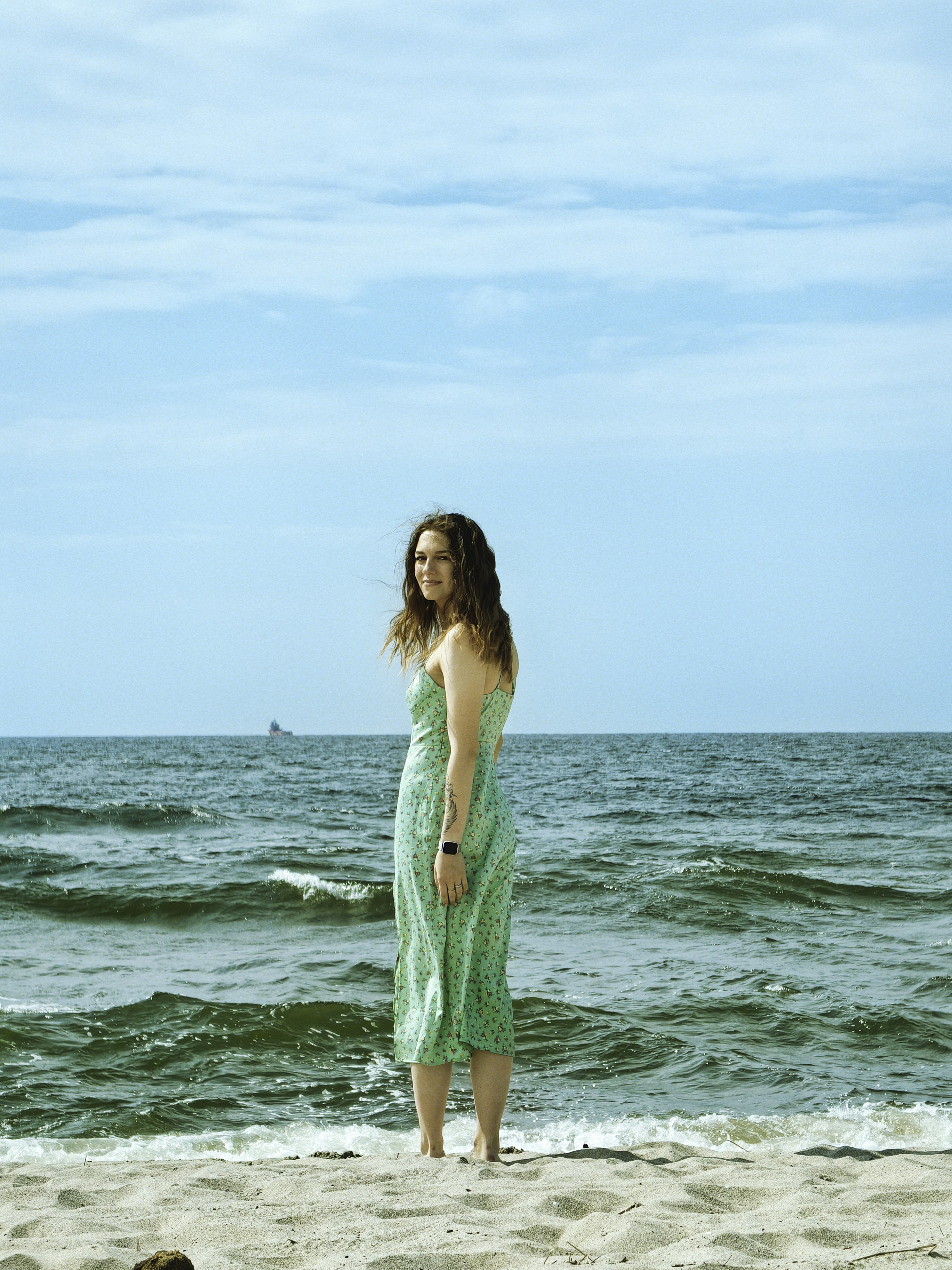 A woman in a light green patterned dress stands barefoot on a sandy beach, with gentle waves rolling in behind her under a clear blue sky.