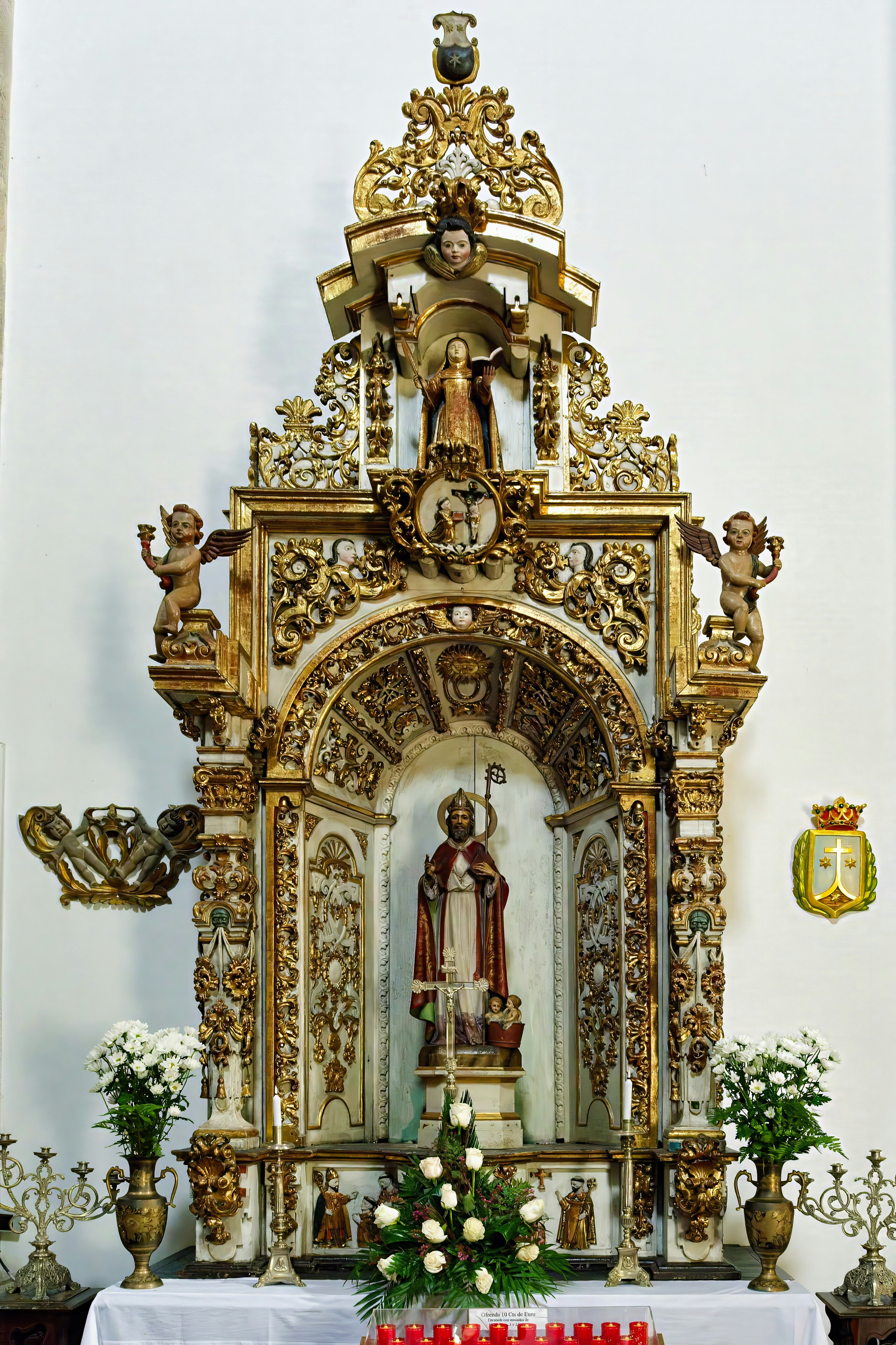 Ornate Altar With Statue of Saint in Church - This image features a large, ornate altar in a church, with a golden and white color scheme. The altar is decorated with intricate carvings and statues, including a central figure of a saint holding a cross. The altar is adorned with white flowers and red candles, adding to the religious and ceremonial atmosphere.