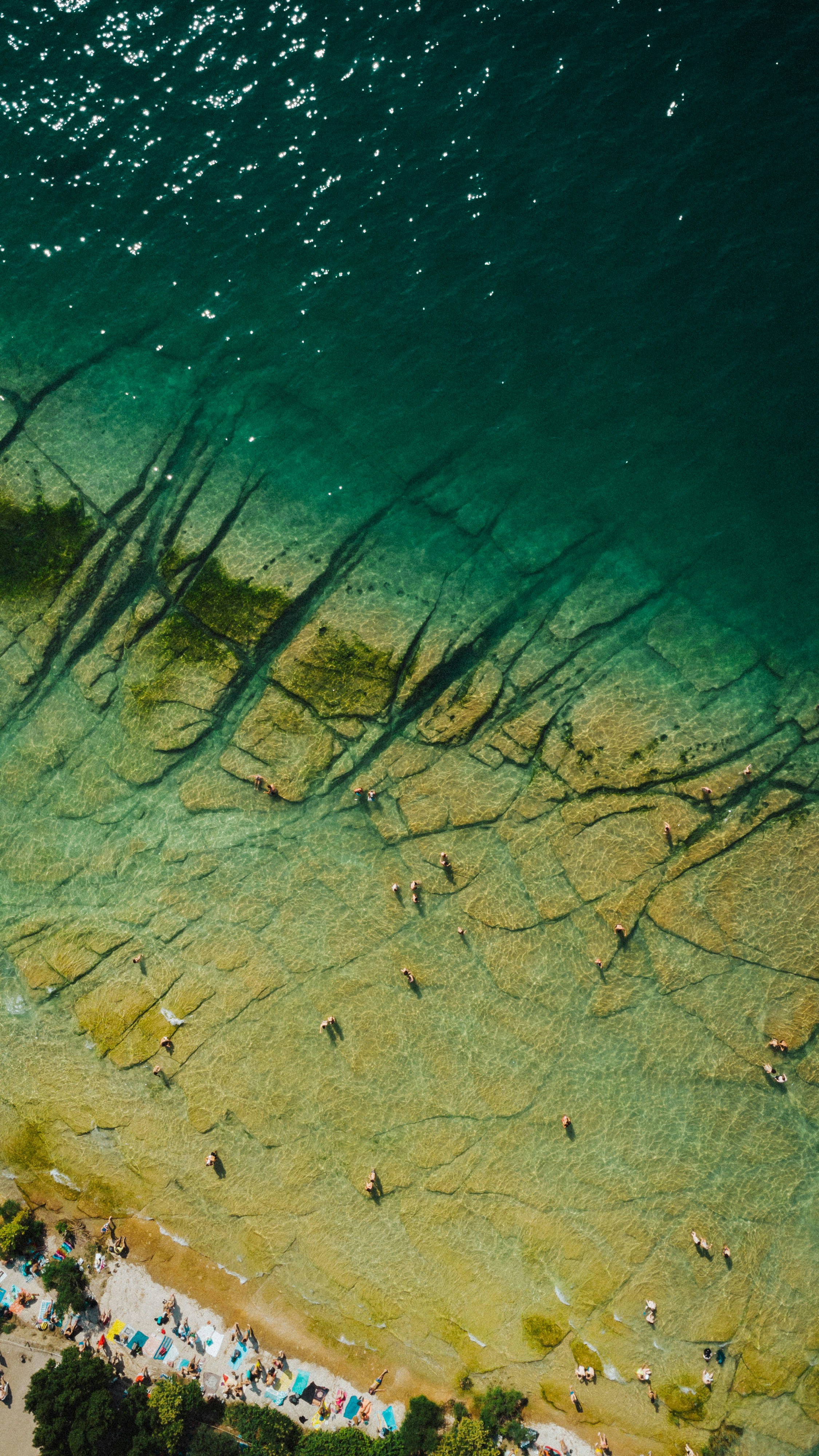 Aerial view of swimmers enjoying a clear, shallow bay with visible rock formations and sandy beach below. Sunlight glimmers on the water's surface.