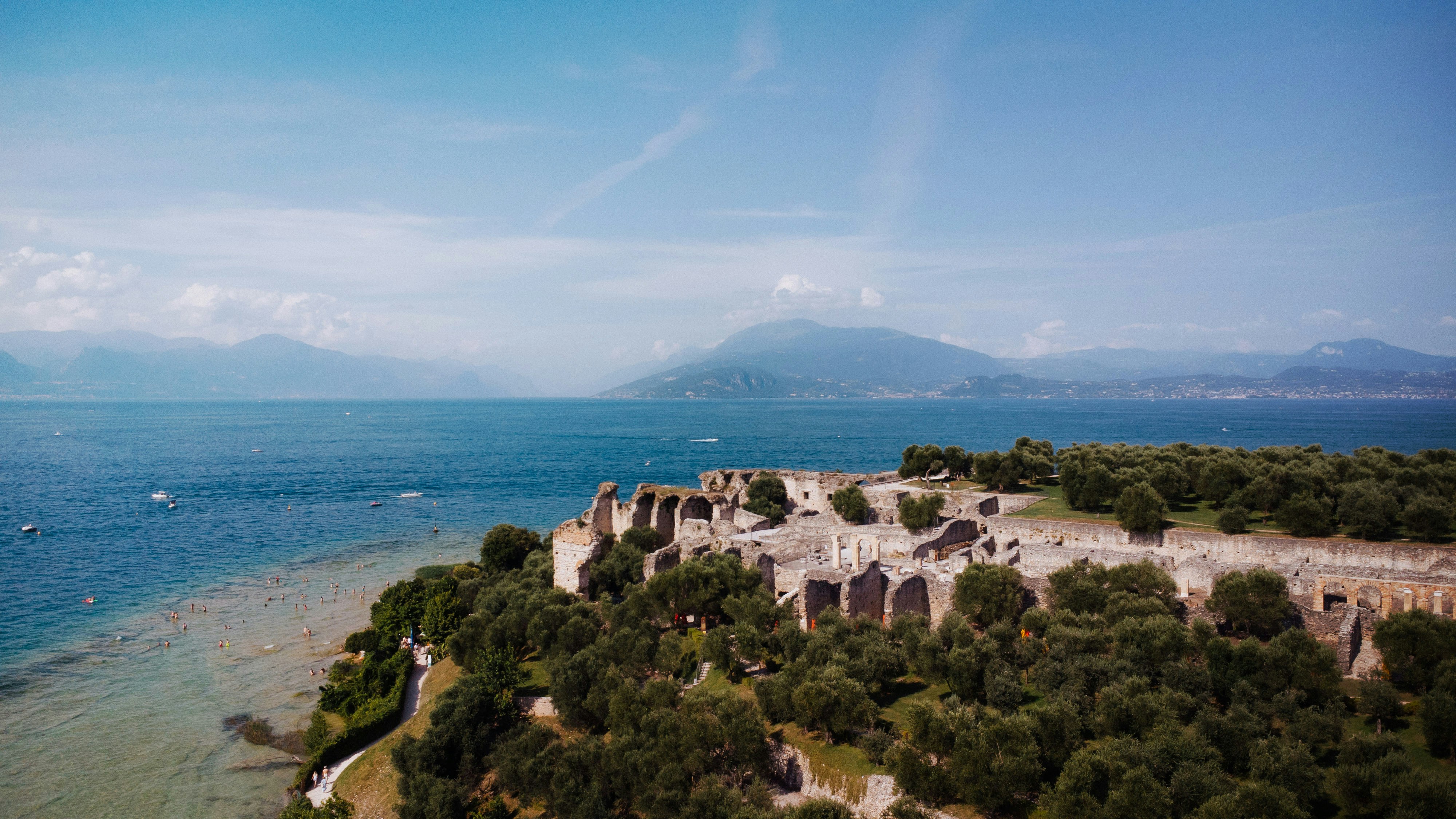 Ancient ruins nestled among lush greenery overlooking a serene lake, with mountains in the distance. The tranquil scene captures a blend of nature and history.