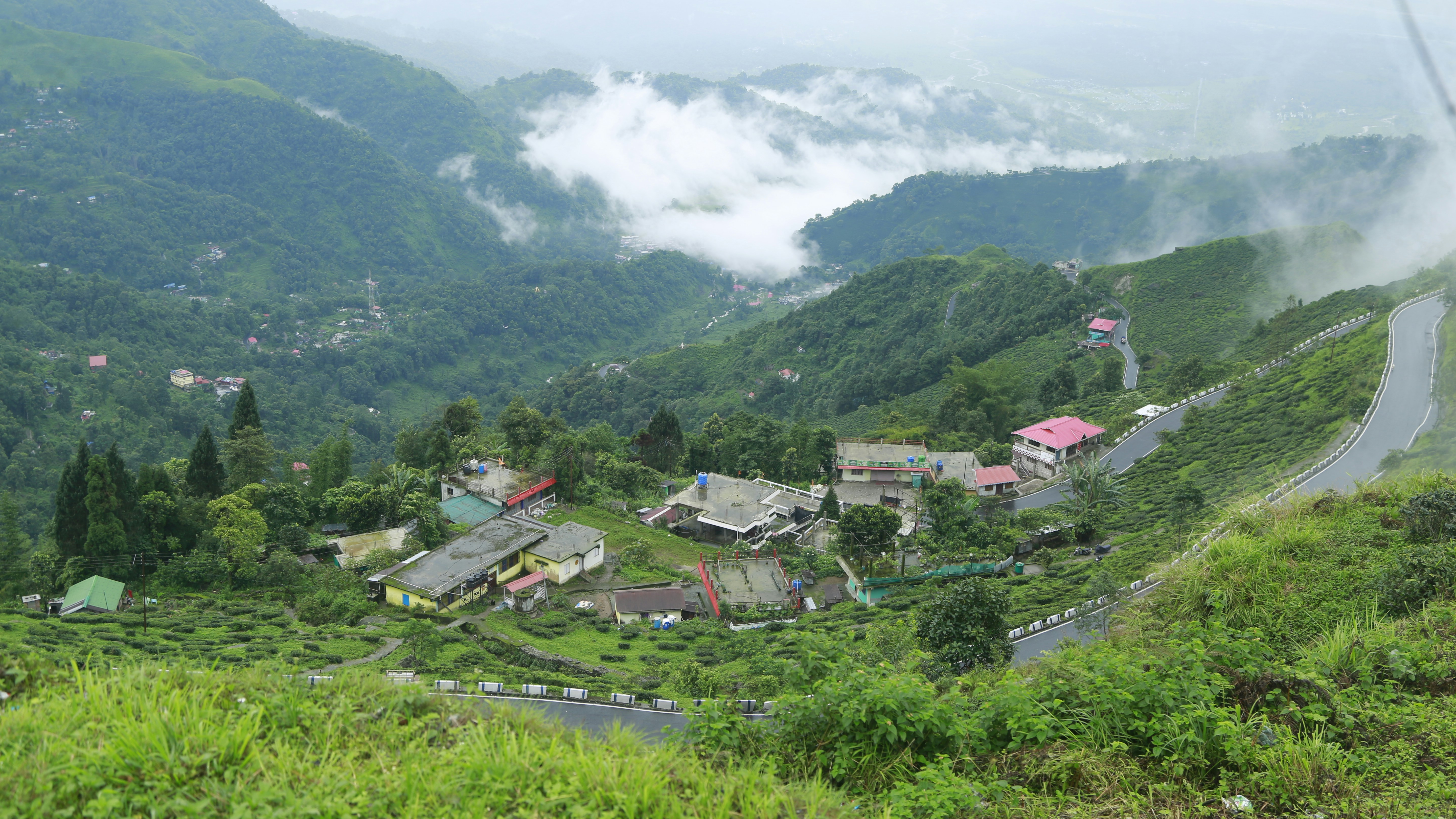 A panoramic view of a quaint mountain village surrounded by lush greenery and rolling hills, shrouded in mist. The winding road leads through the vibrant landscape.