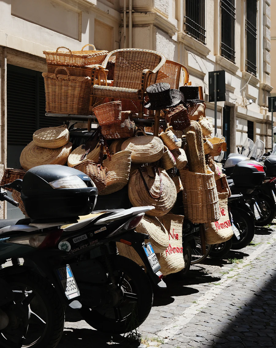 Parked scooters lined along a sunlit street, packed with stacked wicker baskets, hats, and bags. Historic building facades frame the busy, textured scene.