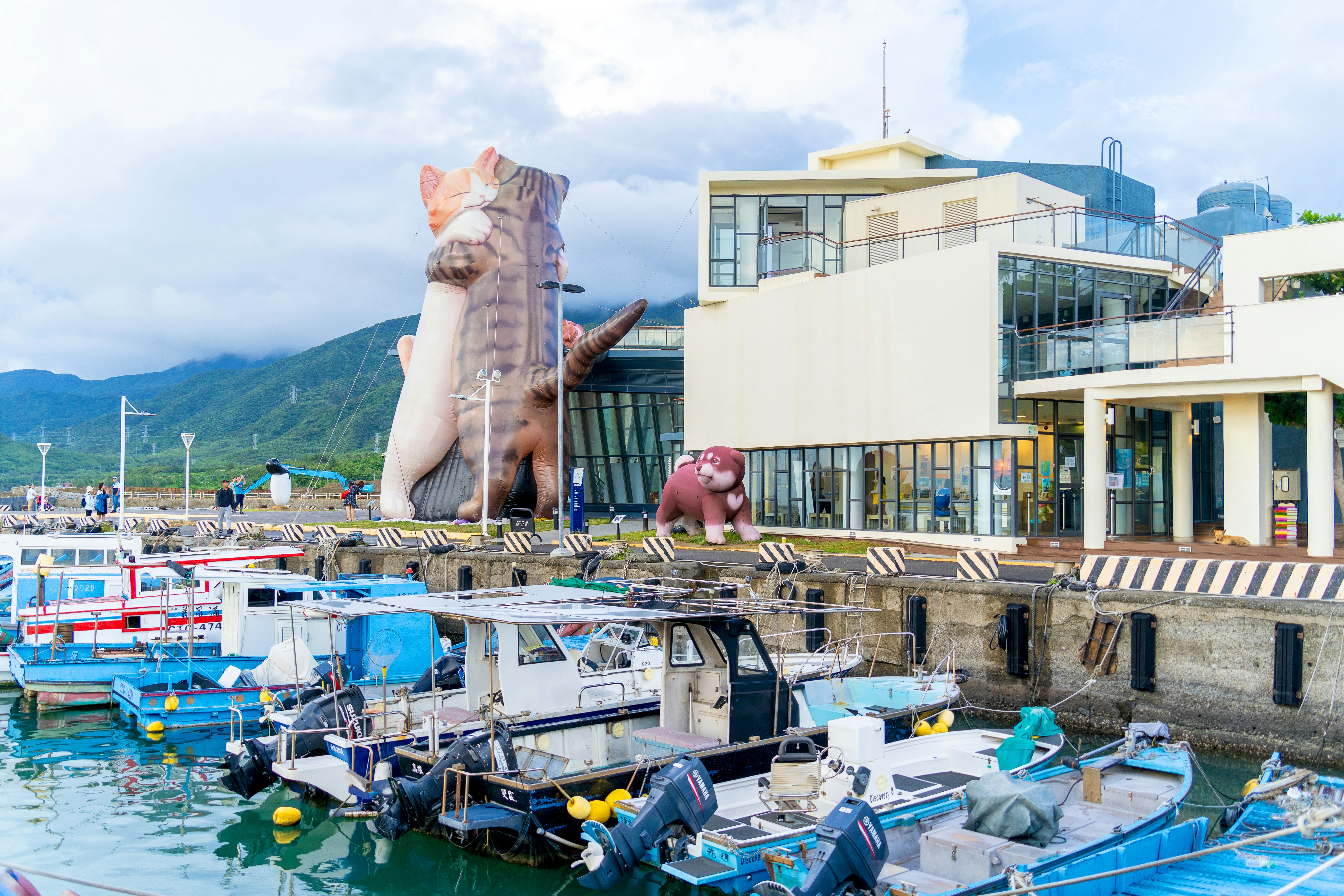 Docked boats beside a modern building with large animal sculptures under a cloudy sky.