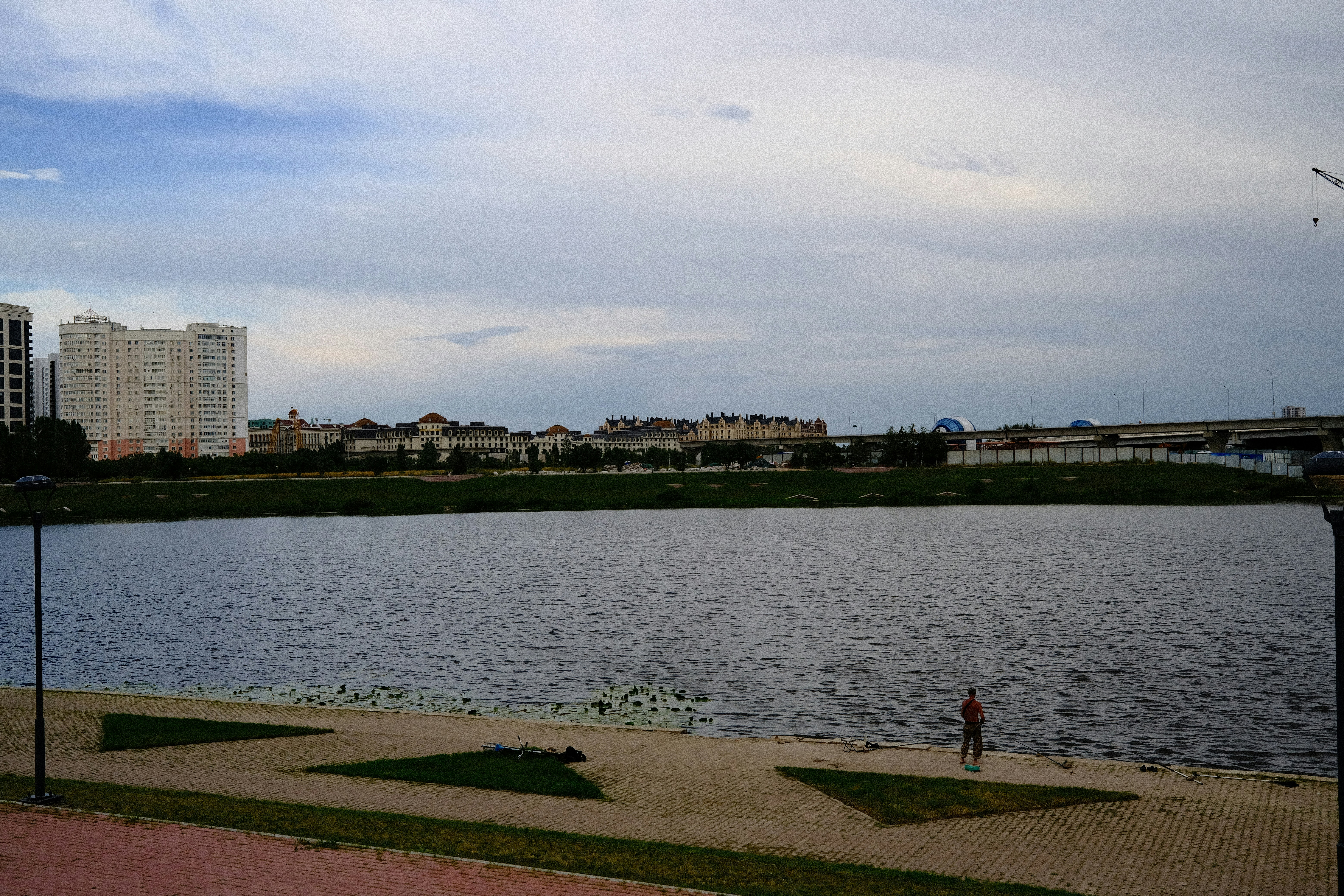 Person walking along a riverside path with cityscape and cloudy sky in the background.