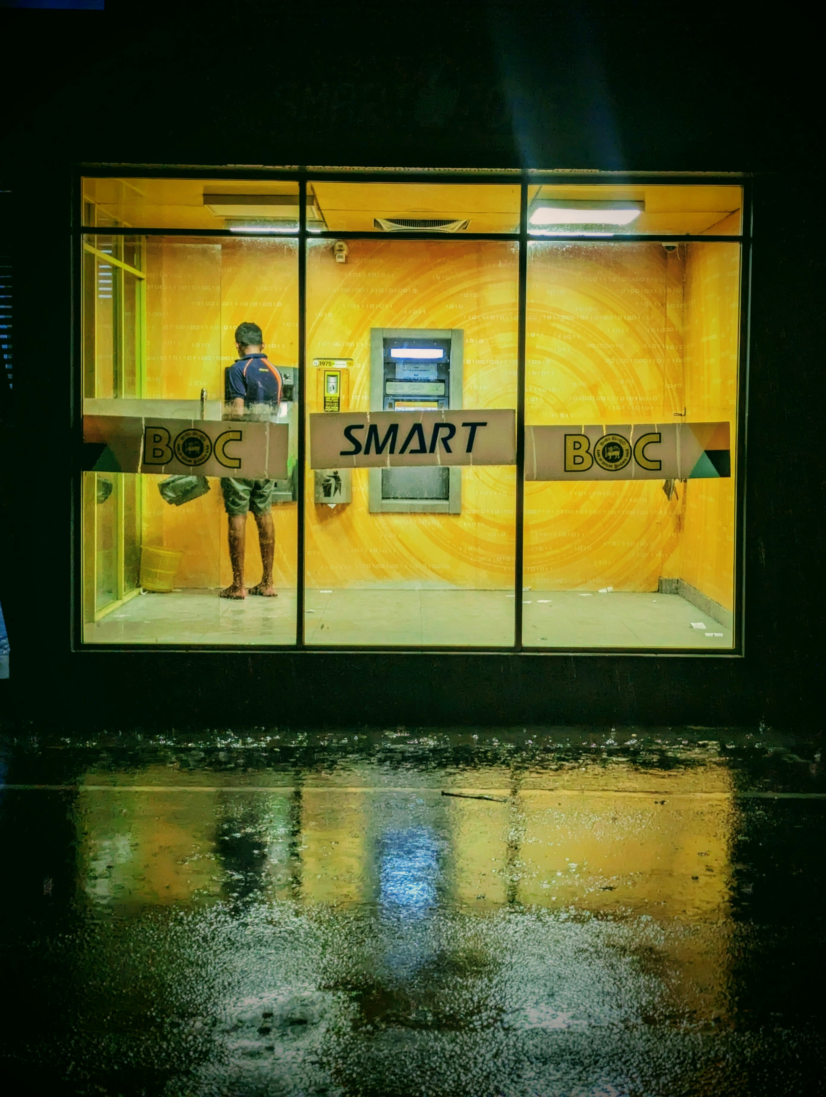 A lone figure stands in a brightly lit, empty phone booth, surrounded by vibrant orange walls and reflections on the wet pavement.