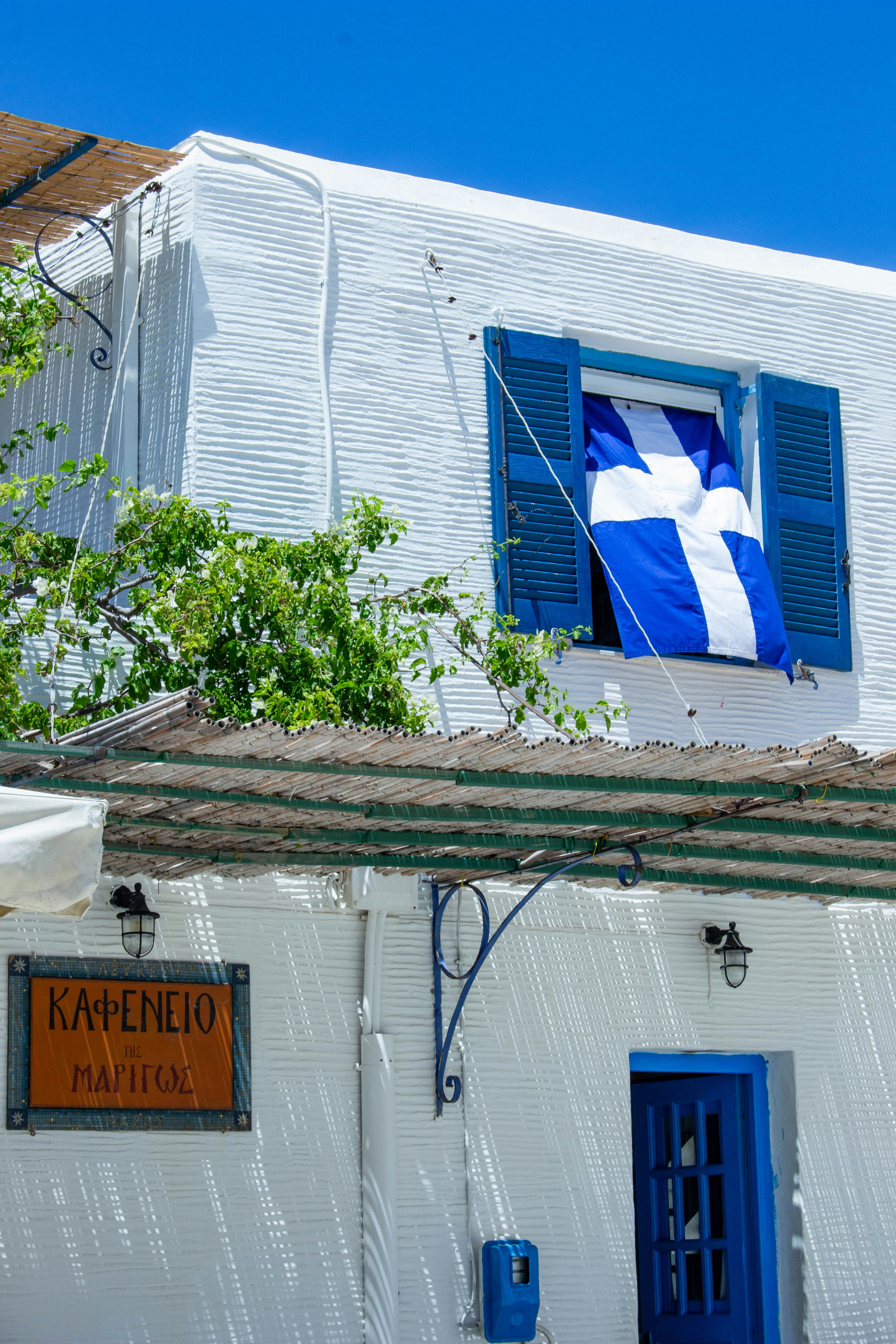 Blue Shutters and a Flag-Draped Window on a Sunlit Greek Café Wall
