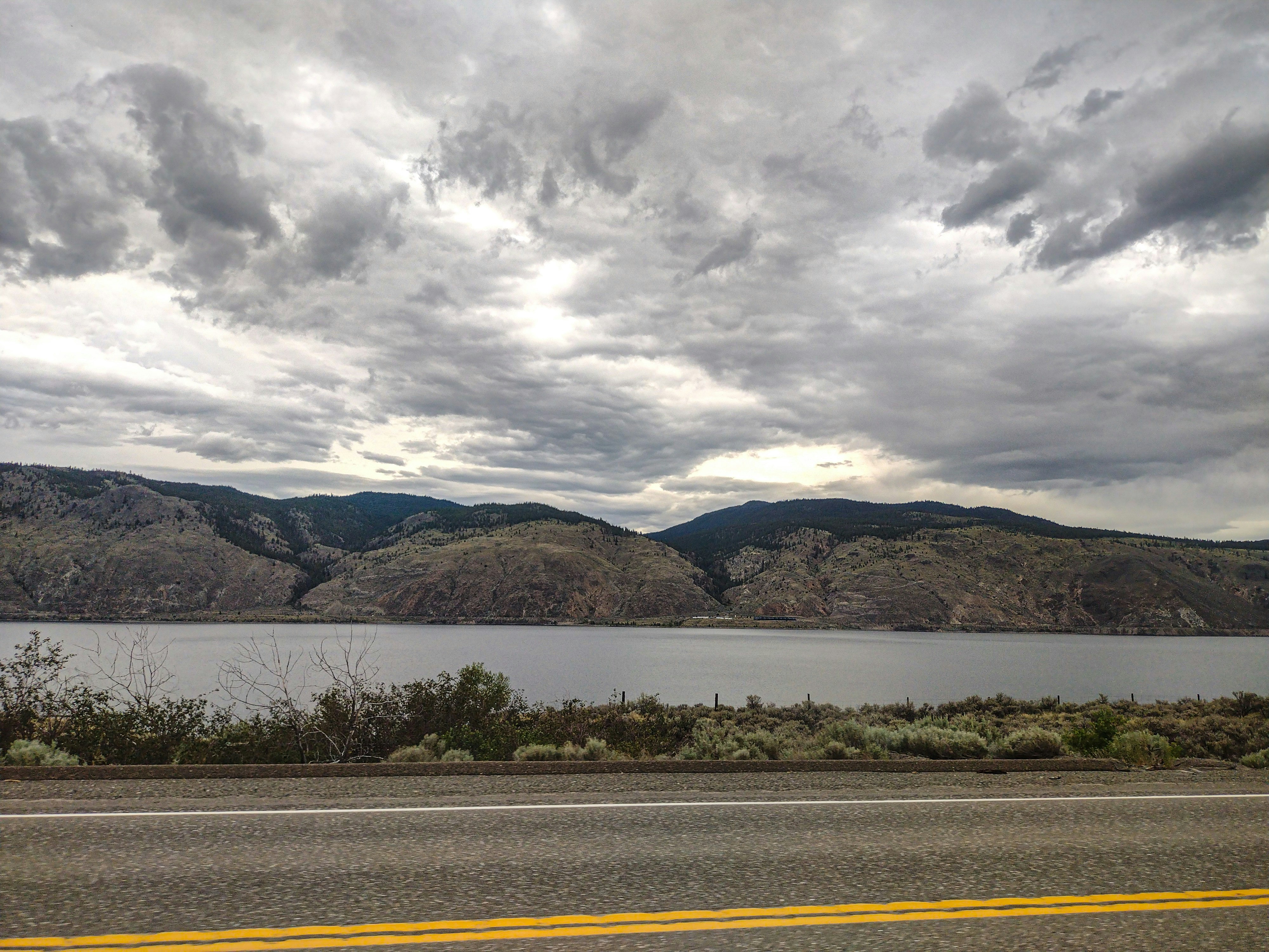 Cloud-laden sky over distant hills and a calm lake beside a road.