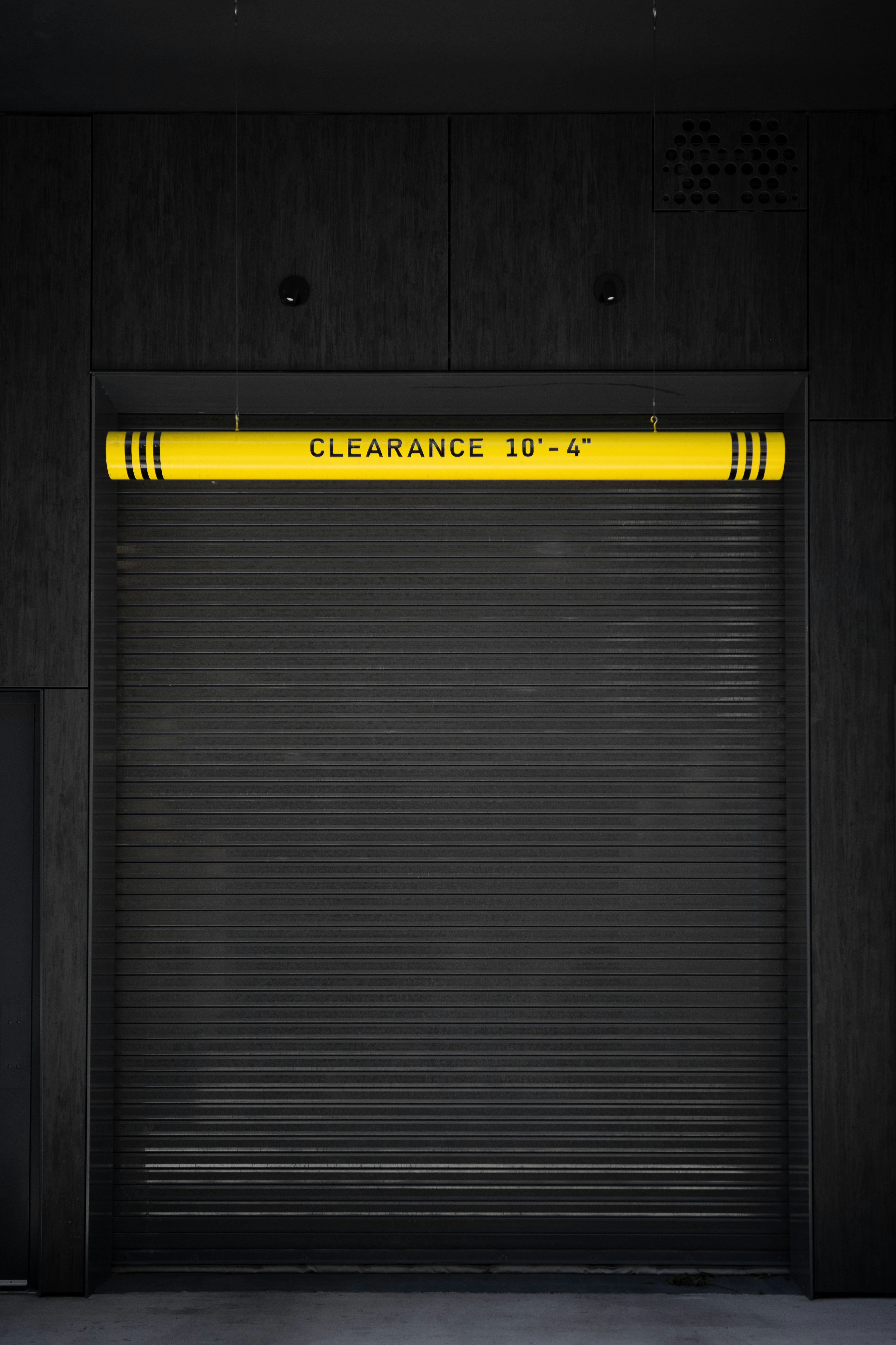Yellow clearance sign displaying 'CLEARANCE 10'-4''' above a dark, corrugated garage door. The stark contrast emphasizes the importance of height restrictions.