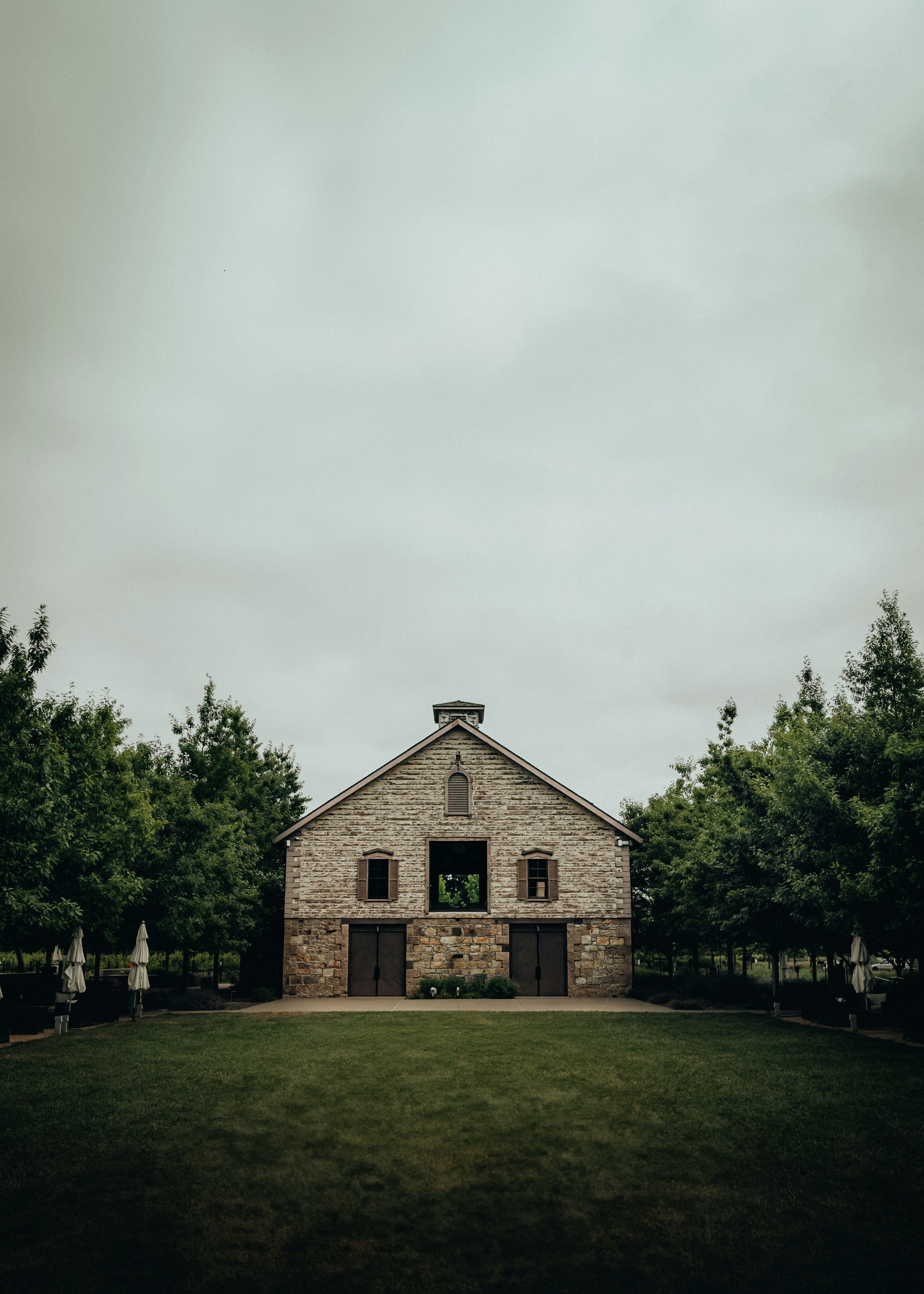 Weathered stone barn framed by lush greenery and shaded seating areas, set against an overcast sky.