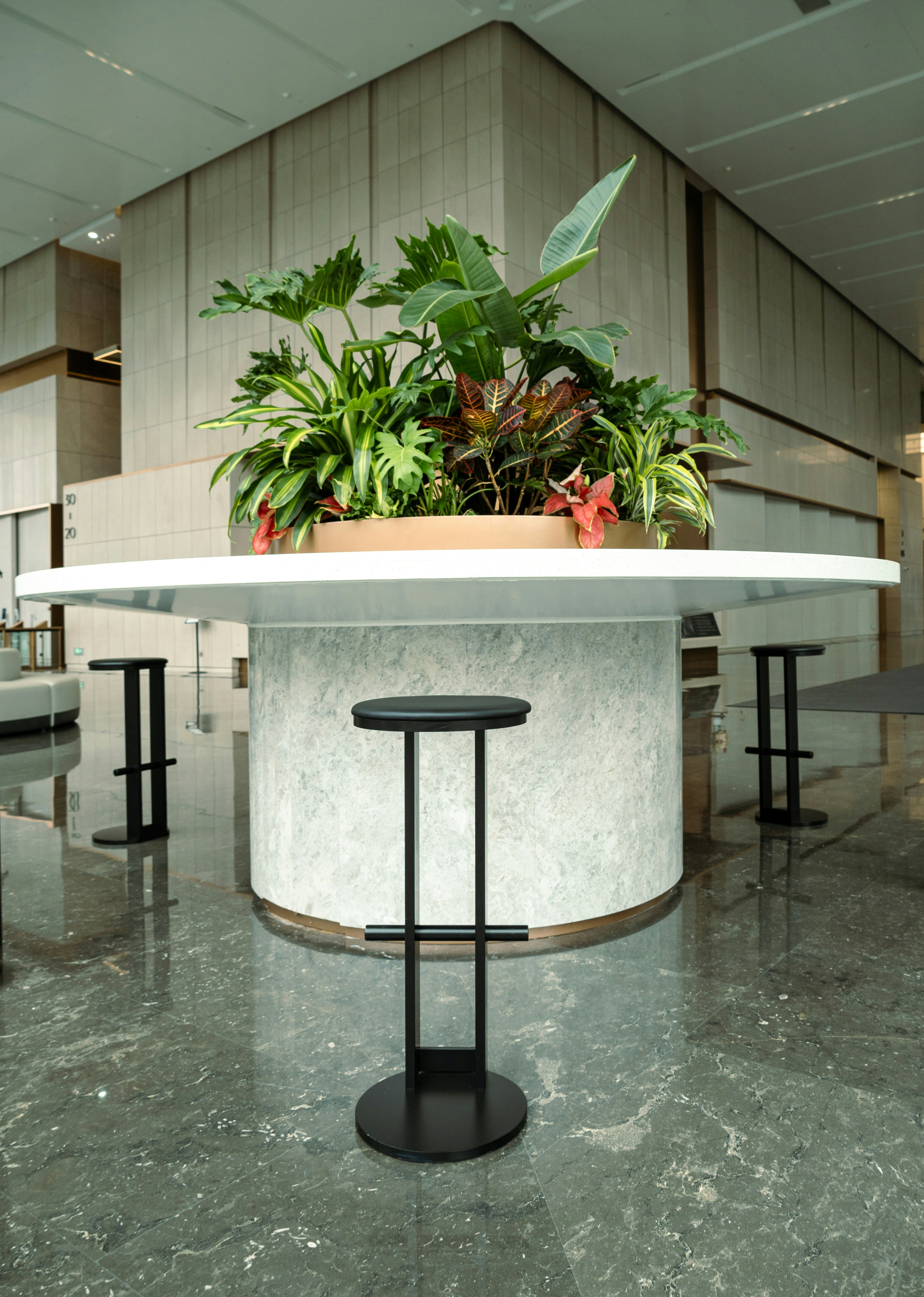 A circular planter brimming with tropical foliage sits atop a marble pedestal in a sleek lobby. Glossy floors and minimalist black stools frame the scene.