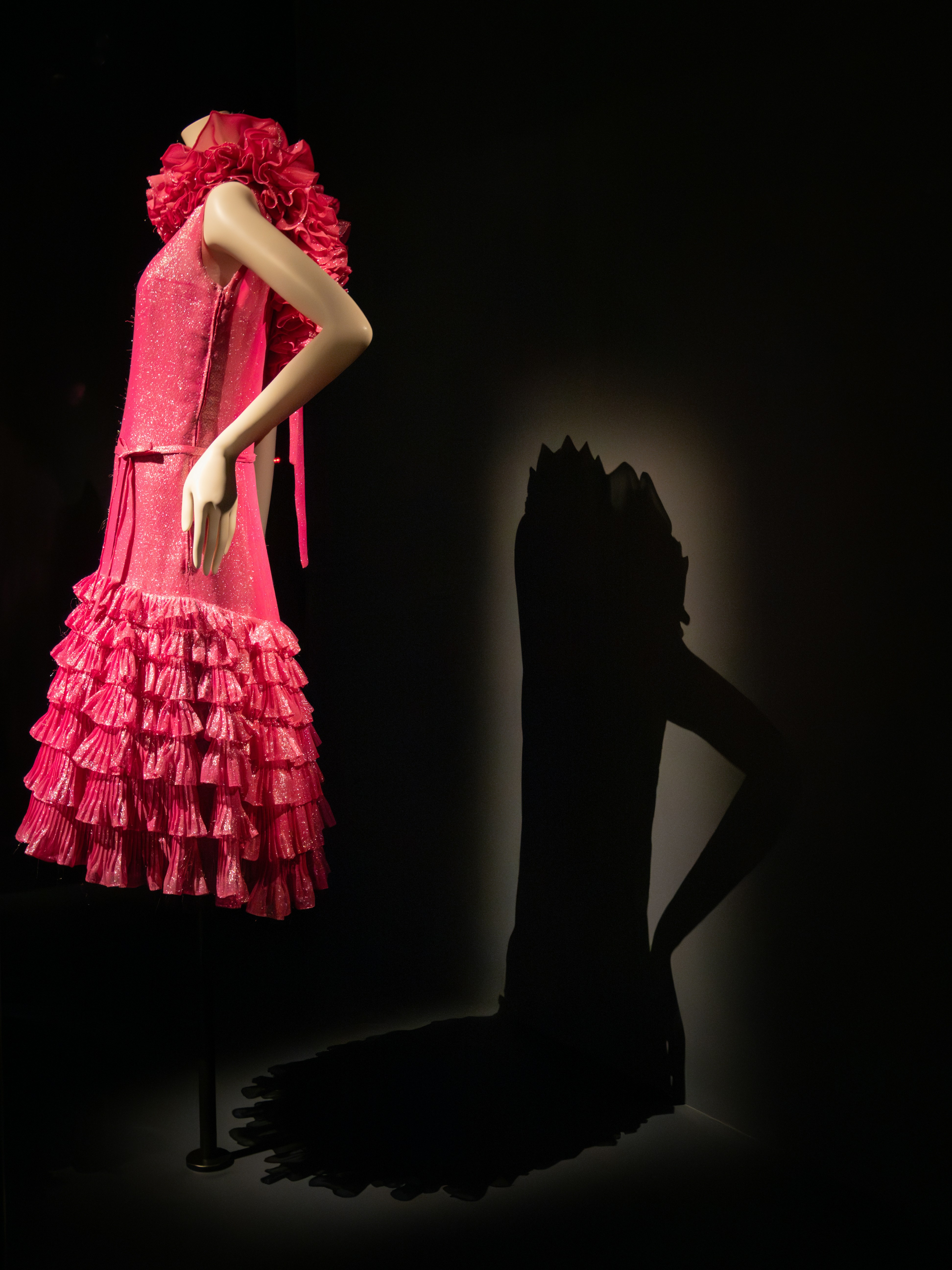 Vibrant pink dress with intricate ruffles displayed on a mannequin, casting a striking shadow against a dark backdrop.