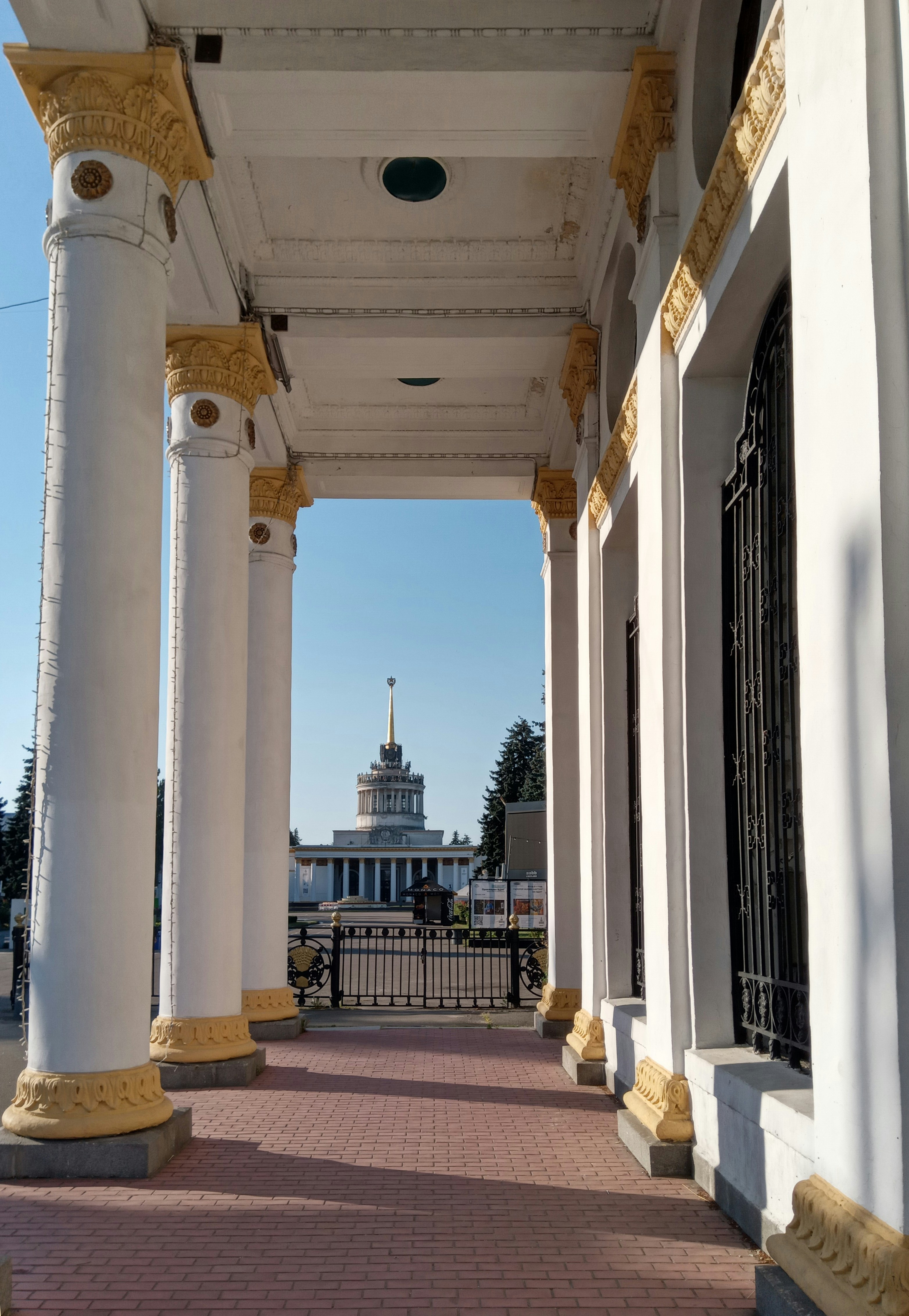 Architectural photograph of a colonnade with gold accents framing a distant city building and spire. The composition emphasizes perspective and symmetry.