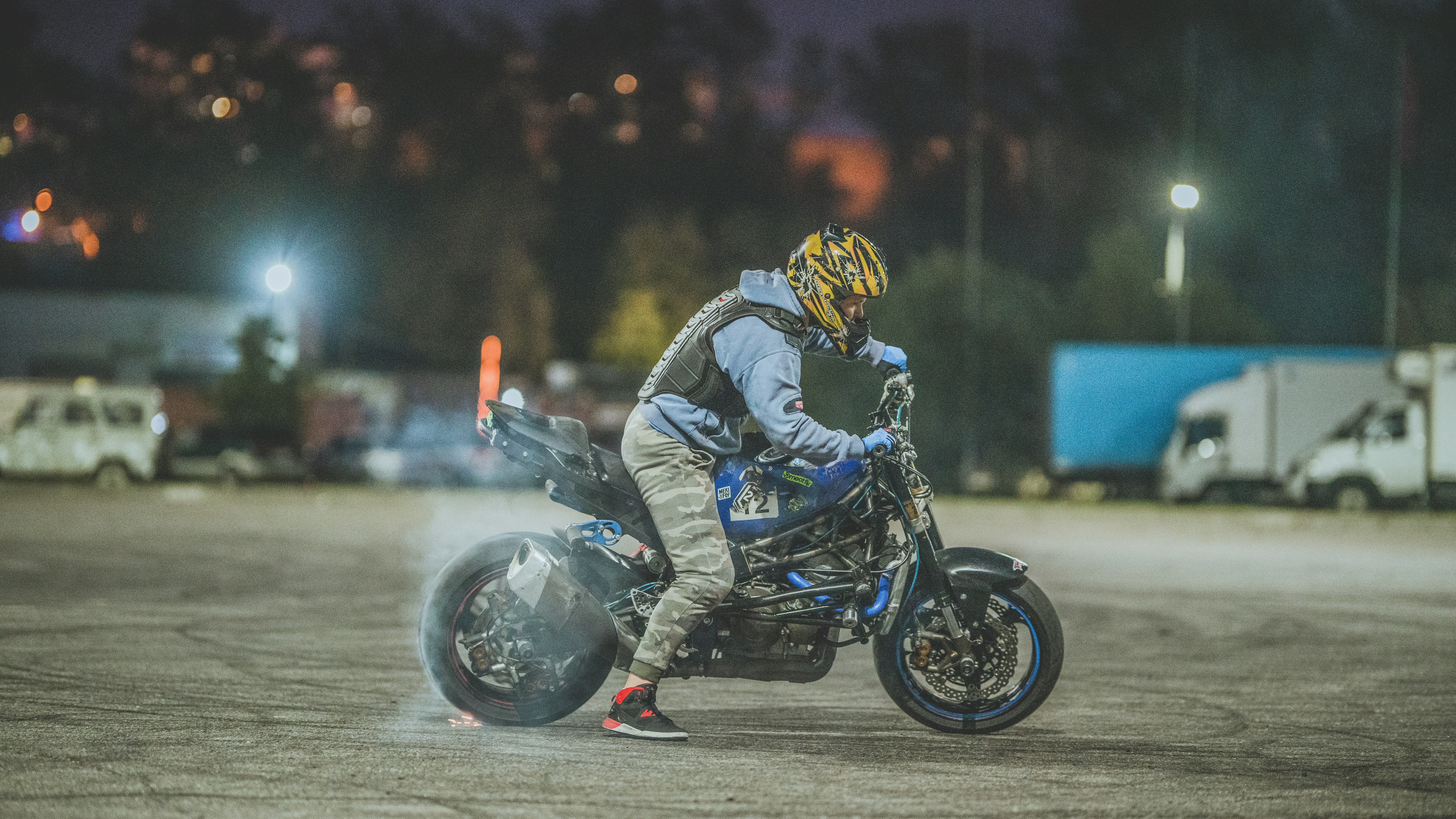 Motorcyclist performing a stunt in an empty parking lot at night, surrounded by soft glowing lights and smoke from the rear tire.