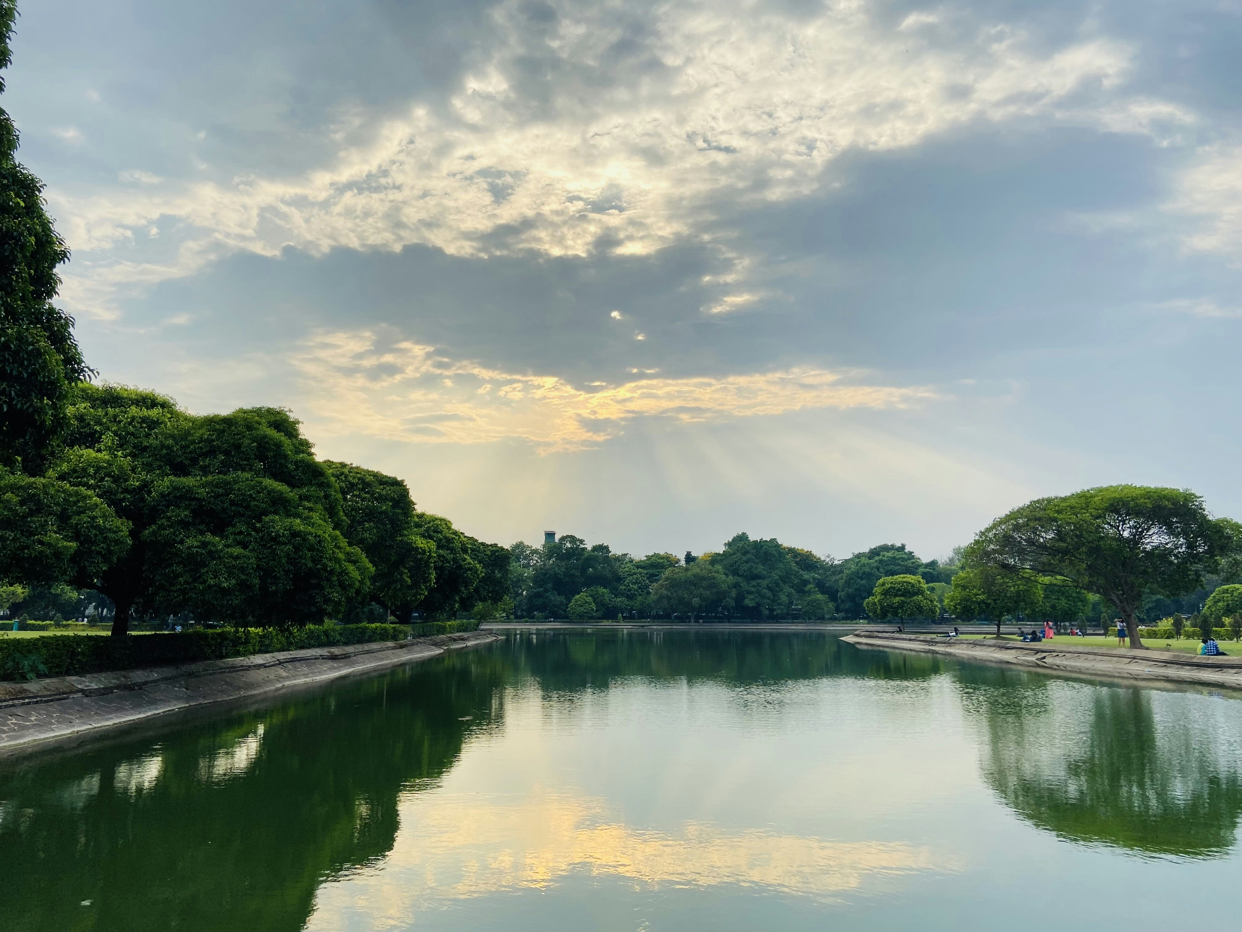 Serene lake reflecting lush trees and dramatic cloud formations with sunbeams breaking through.