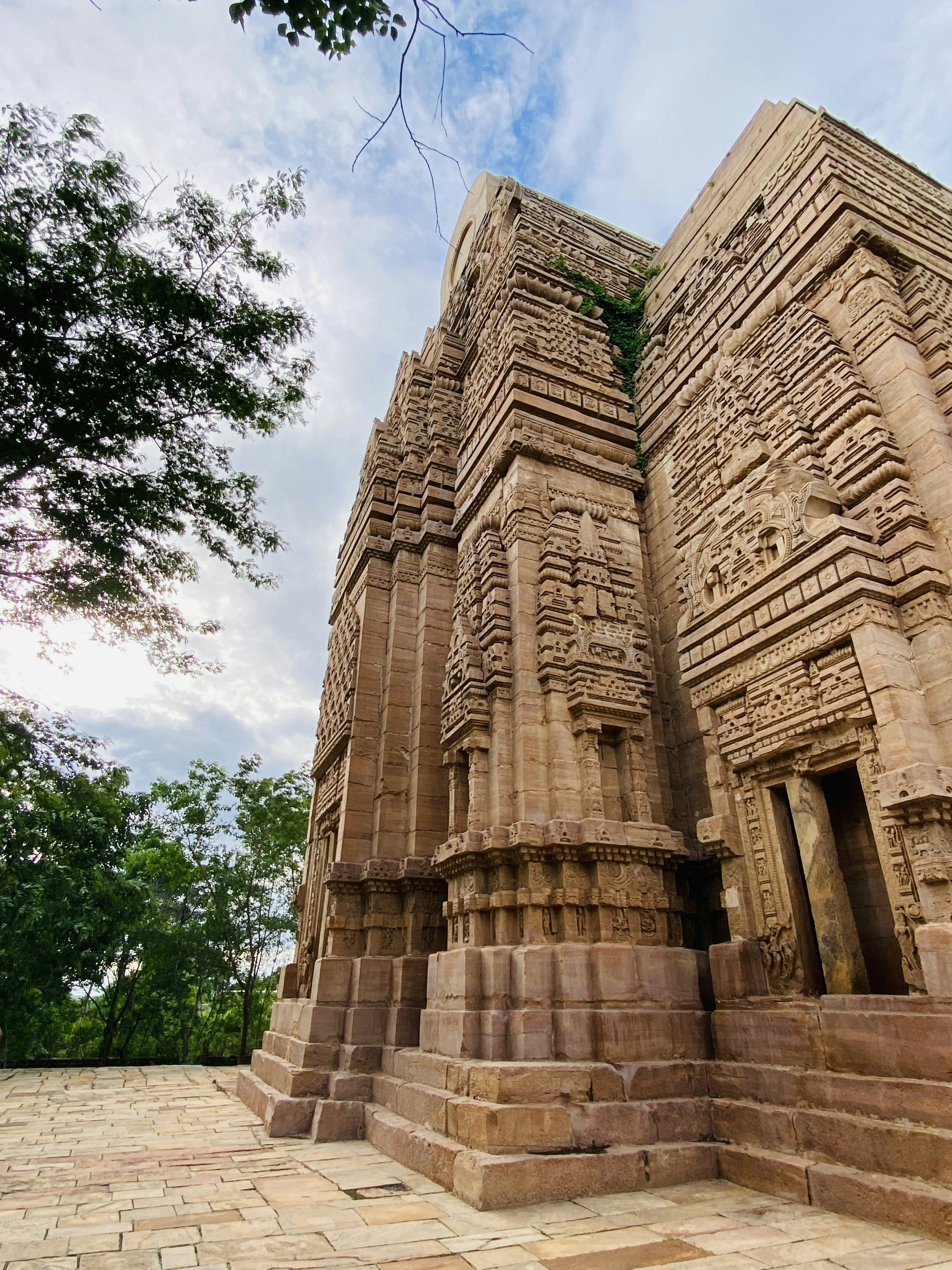 Elaborately carved stone temple facade rising against a backdrop of lush trees and a partly cloudy sky.