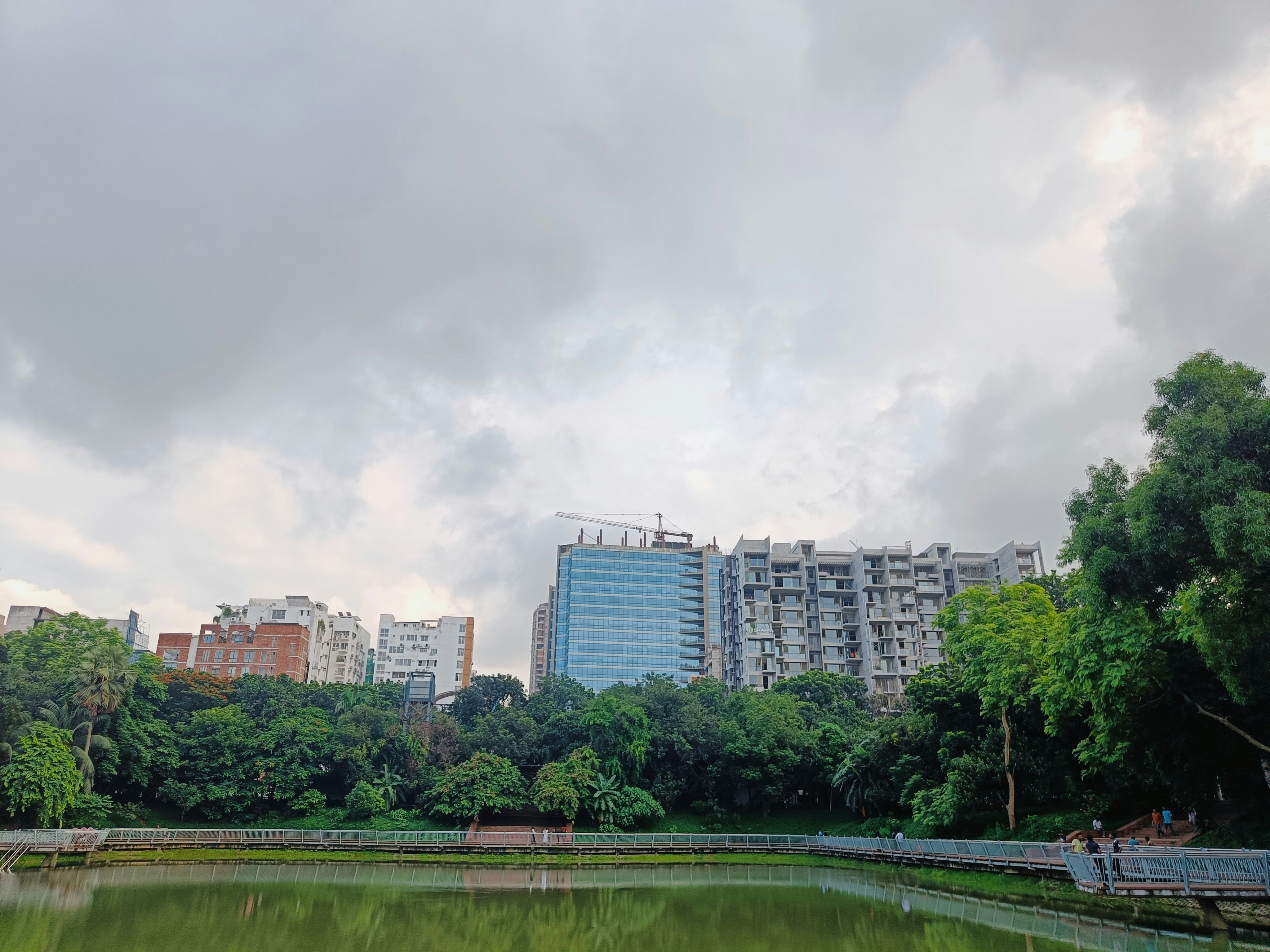 In this captivating scene, nature and urban life converge harmoniously. A serene lake reflects the azure sky overhead, framed by lush, vibrant greenery of a dense forest. Amidst this natural beauty, a cluster of tall buildings rises in the distance, standing as symbols of human ingenuity against the backdrop of tranquil wilderness. This photograph captures the juxtaposition of urban development and untouched nature, offering a poignant glimpse into the balance between modernity and the environment."