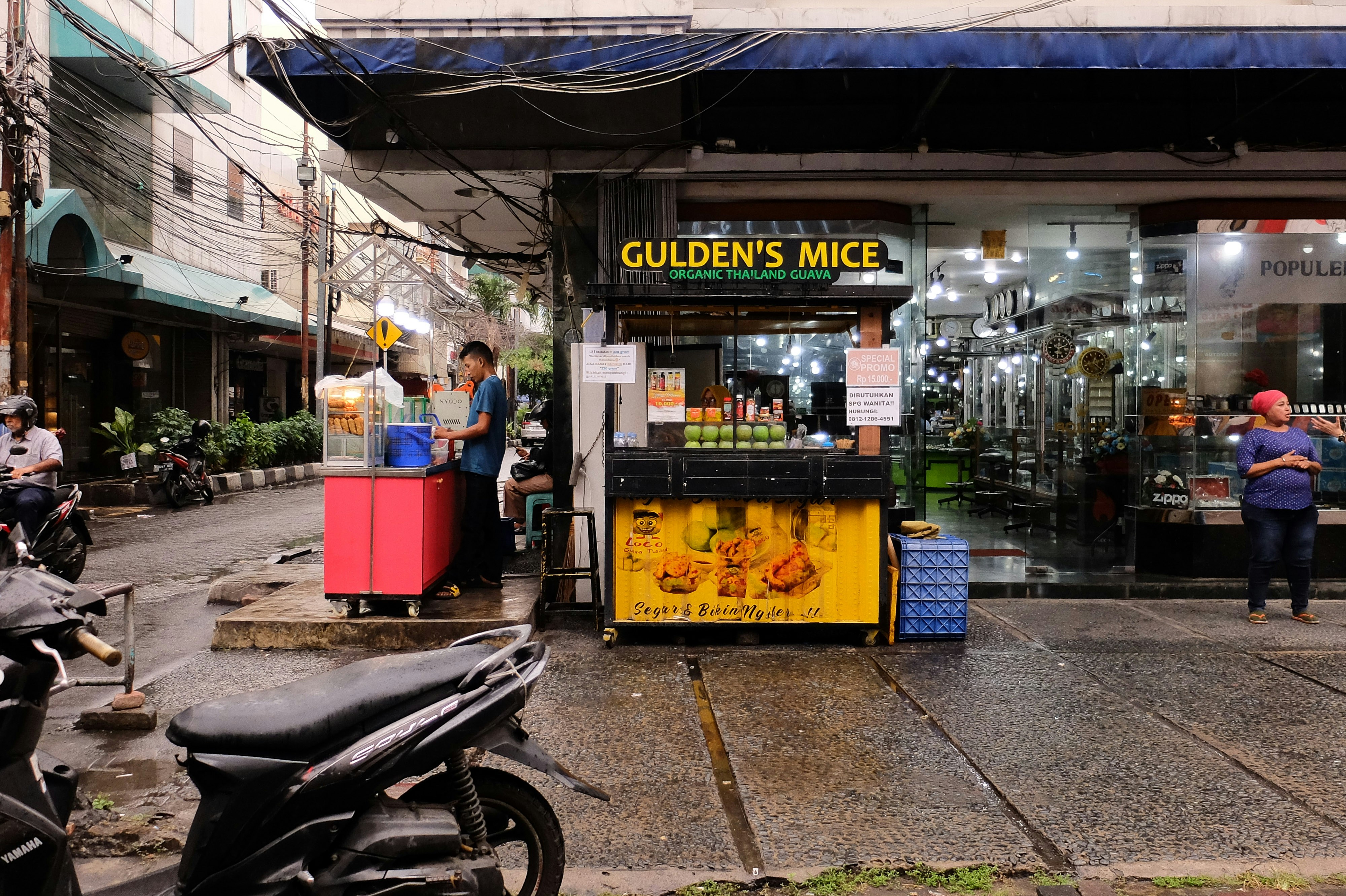 Photograph of a rain-soaked street with a bright yellow-lit food stall at the center. A scooter sits in the foreground while pedestrians browse the market in the background.