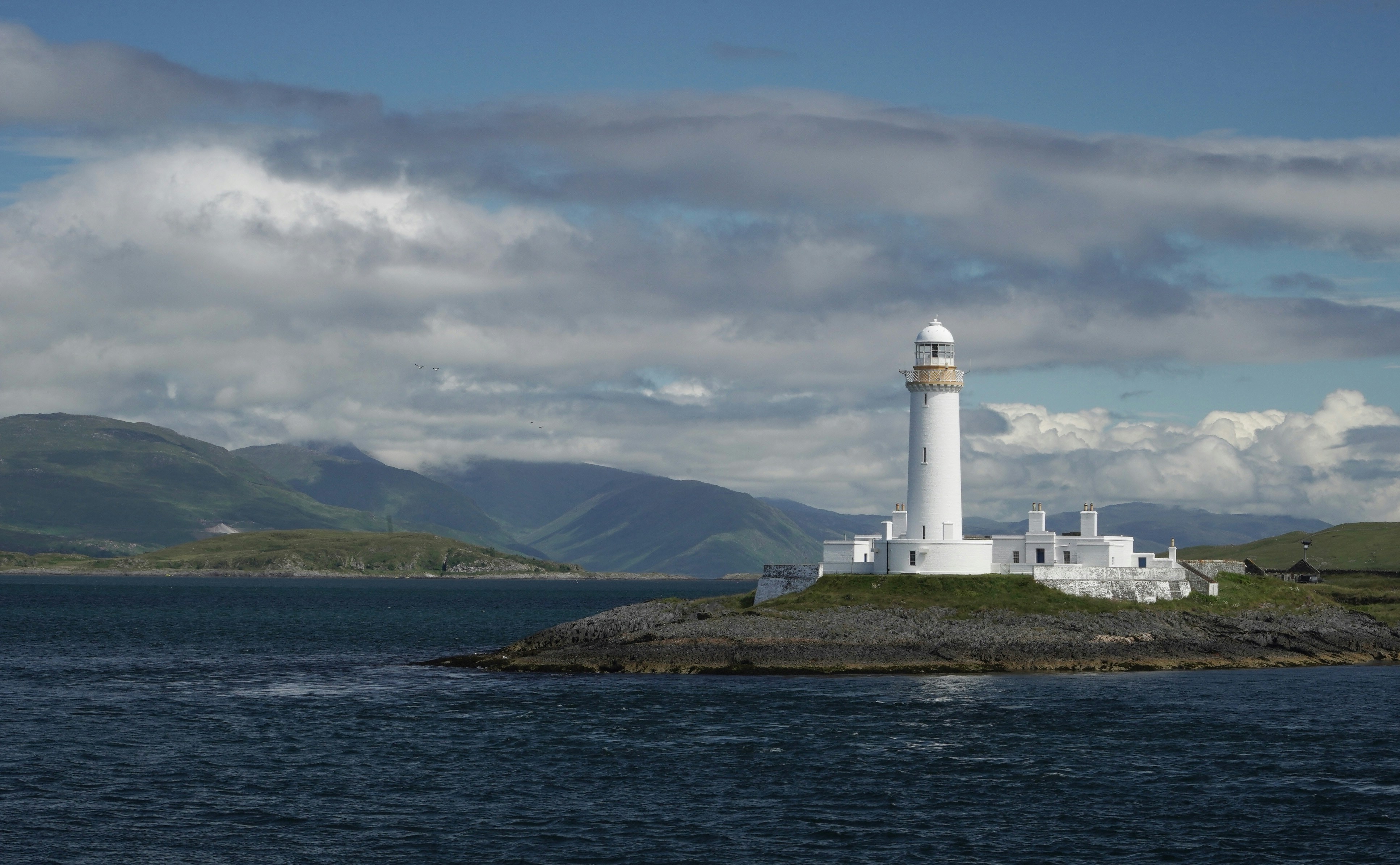 White lighthouse standing on a rocky island with rolling hills in the background under a bright blue sky.