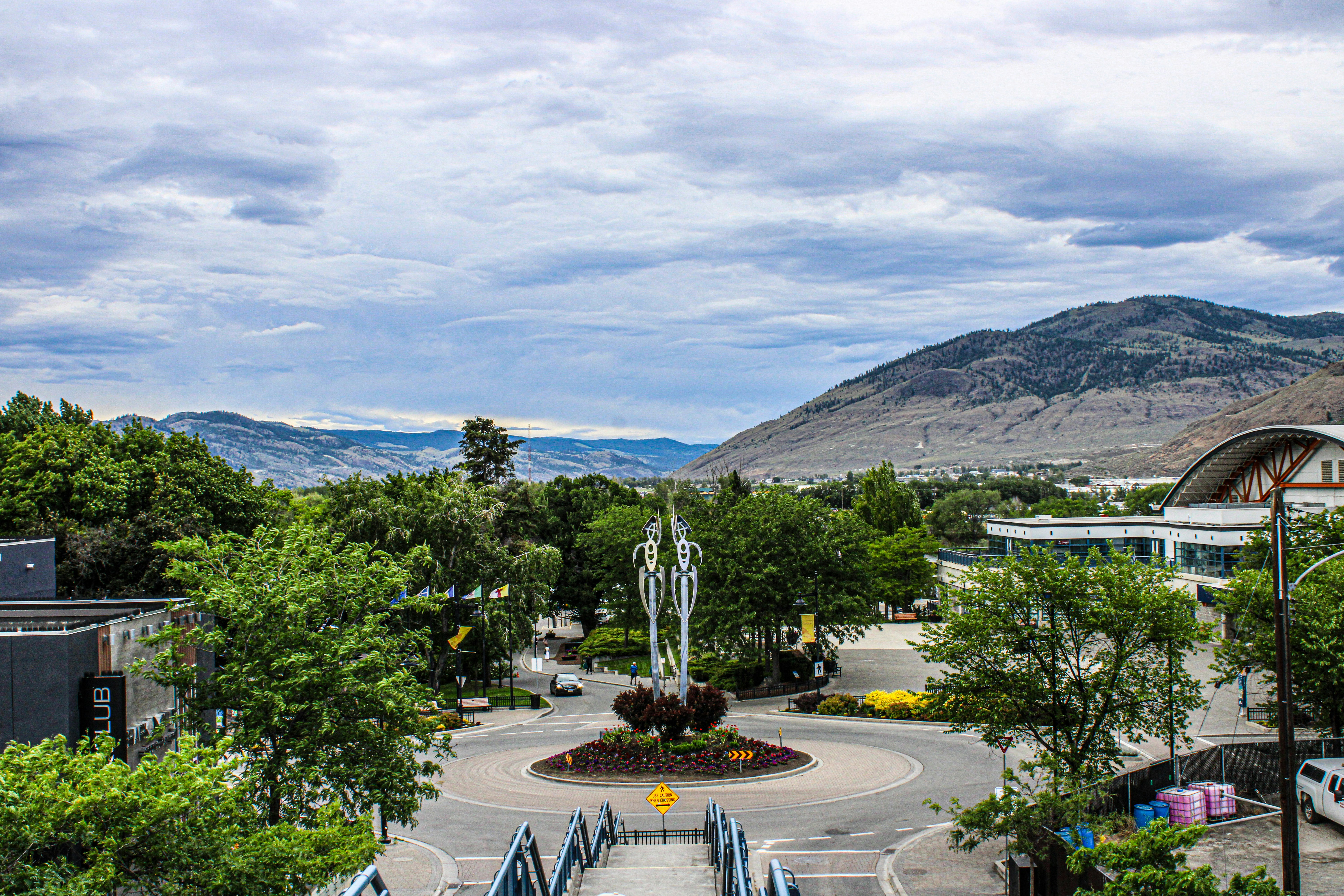 Cityscape with a central statue, surrounded by lush greenery and distant mountains under a cloudy sky.