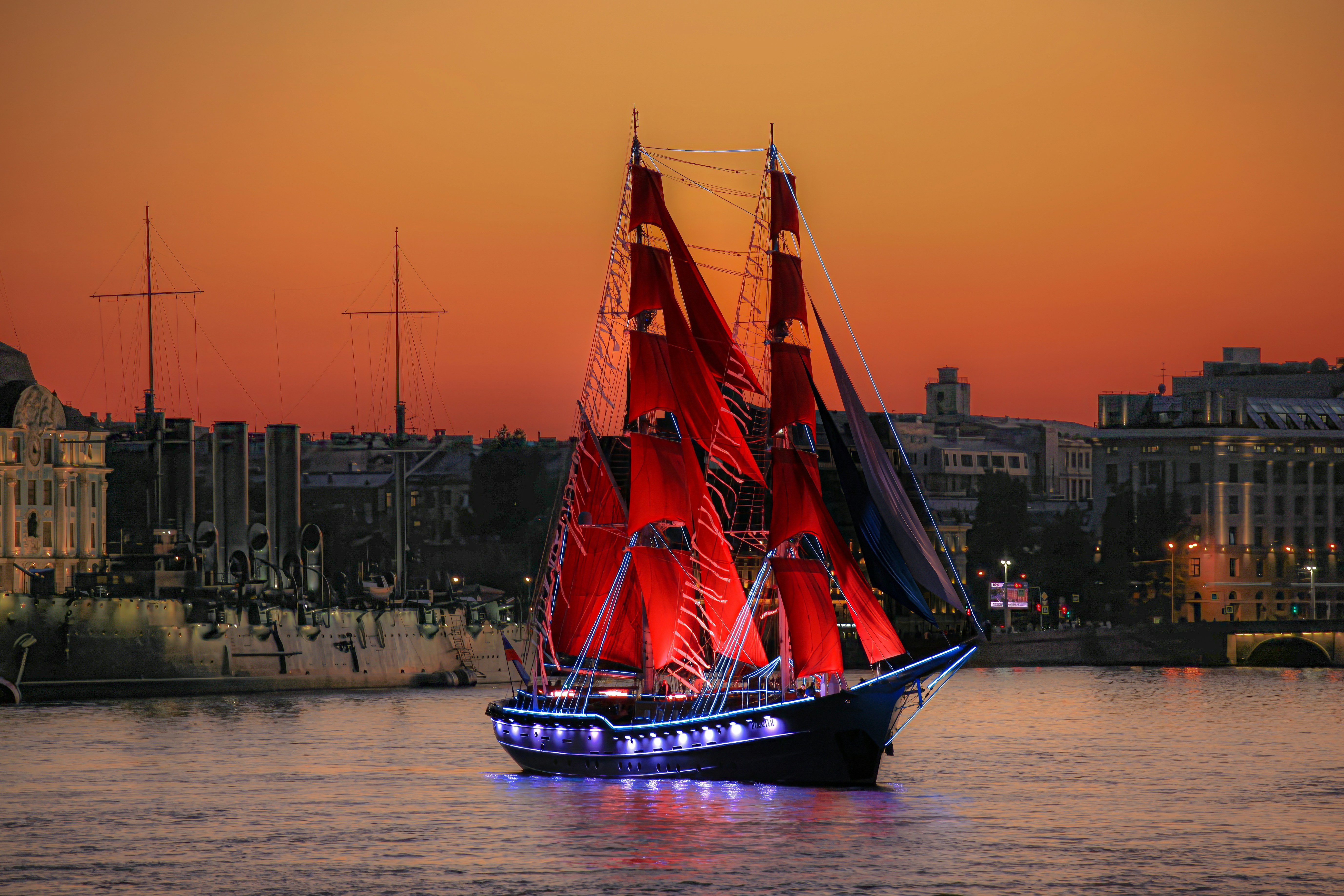 Red-sailed river ship gliding past a historic embankment at sunset