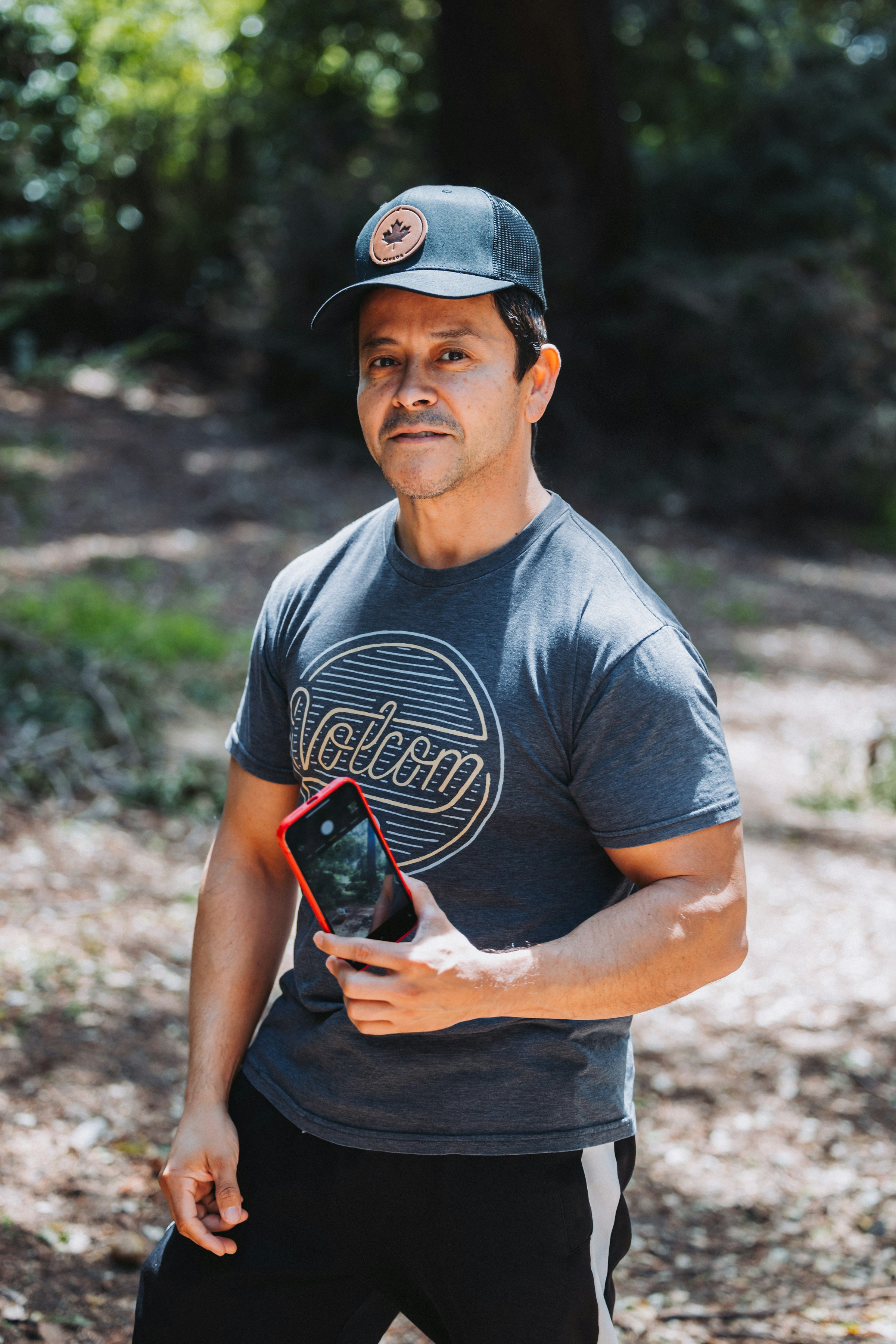 Dappled Light Portrait: A cap-clad man holds a red smartphone in a sunlit woodland