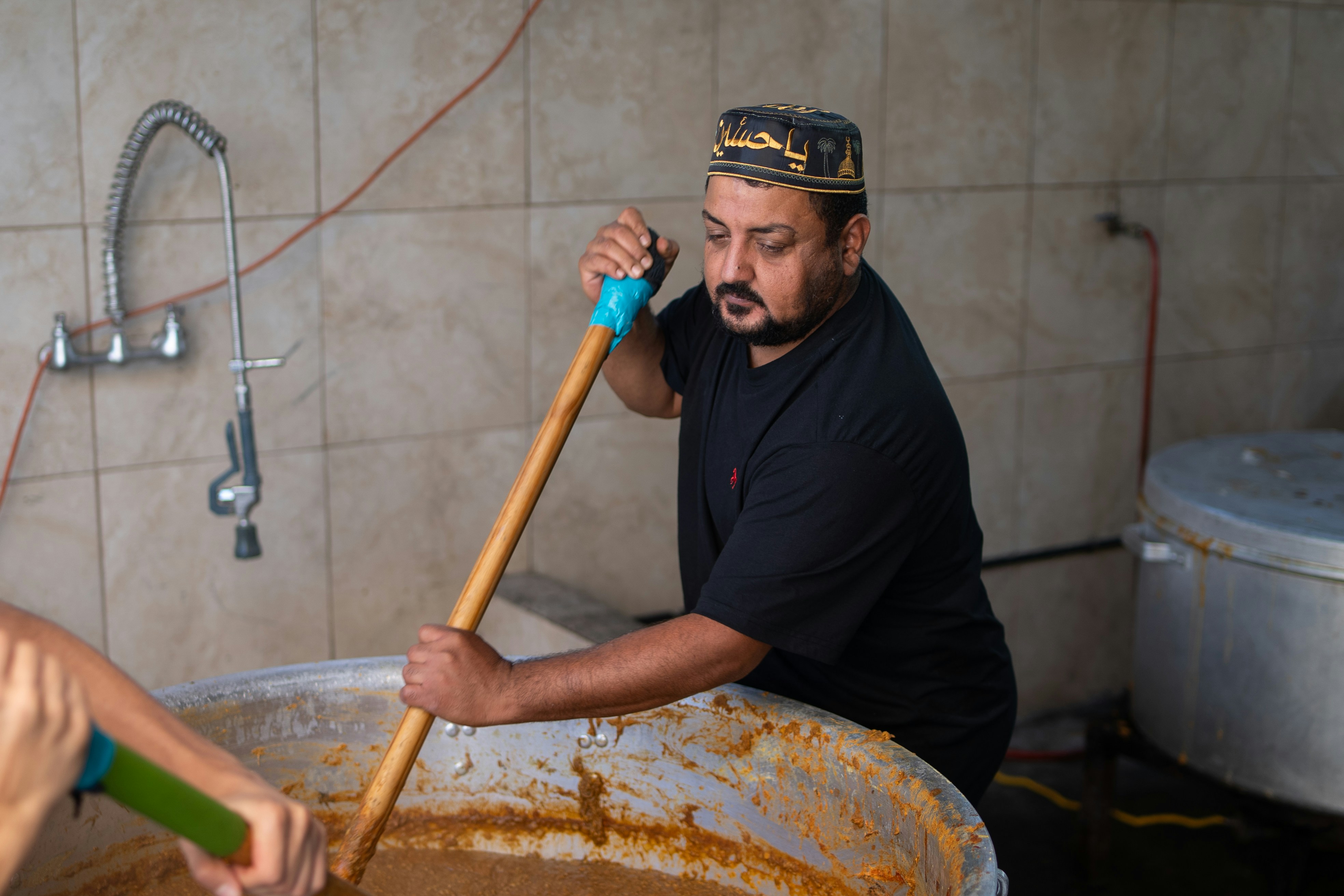 A chef skillfully stirs a large pot of simmering food in a bustling kitchen, showcasing culinary craftsmanship and dedication.