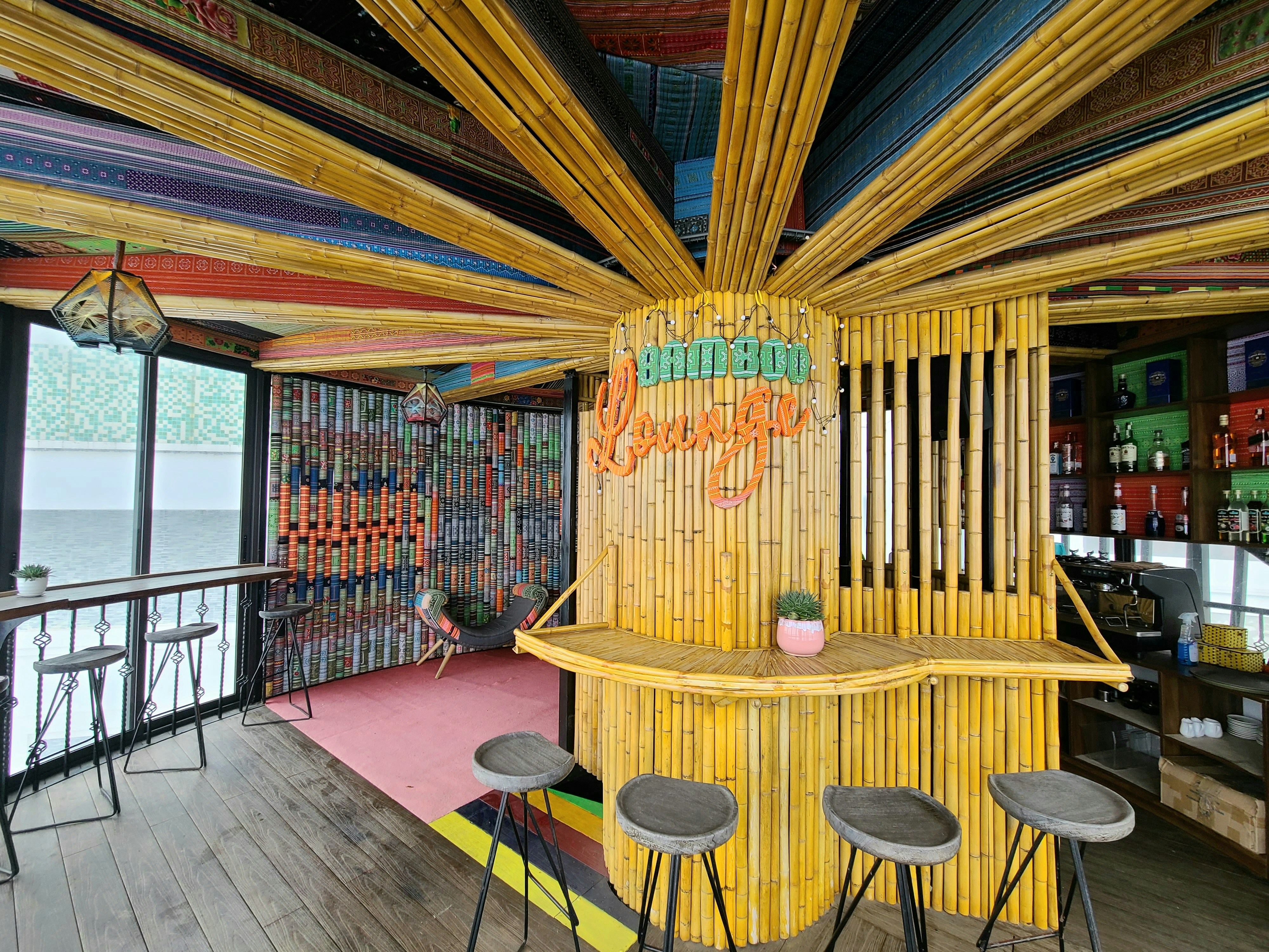 Interior photo of a bamboo-bar lounge featuring a curved counter and gray stools. A neon-style logo sits on the central column, with colorful ceiling beams overhead.