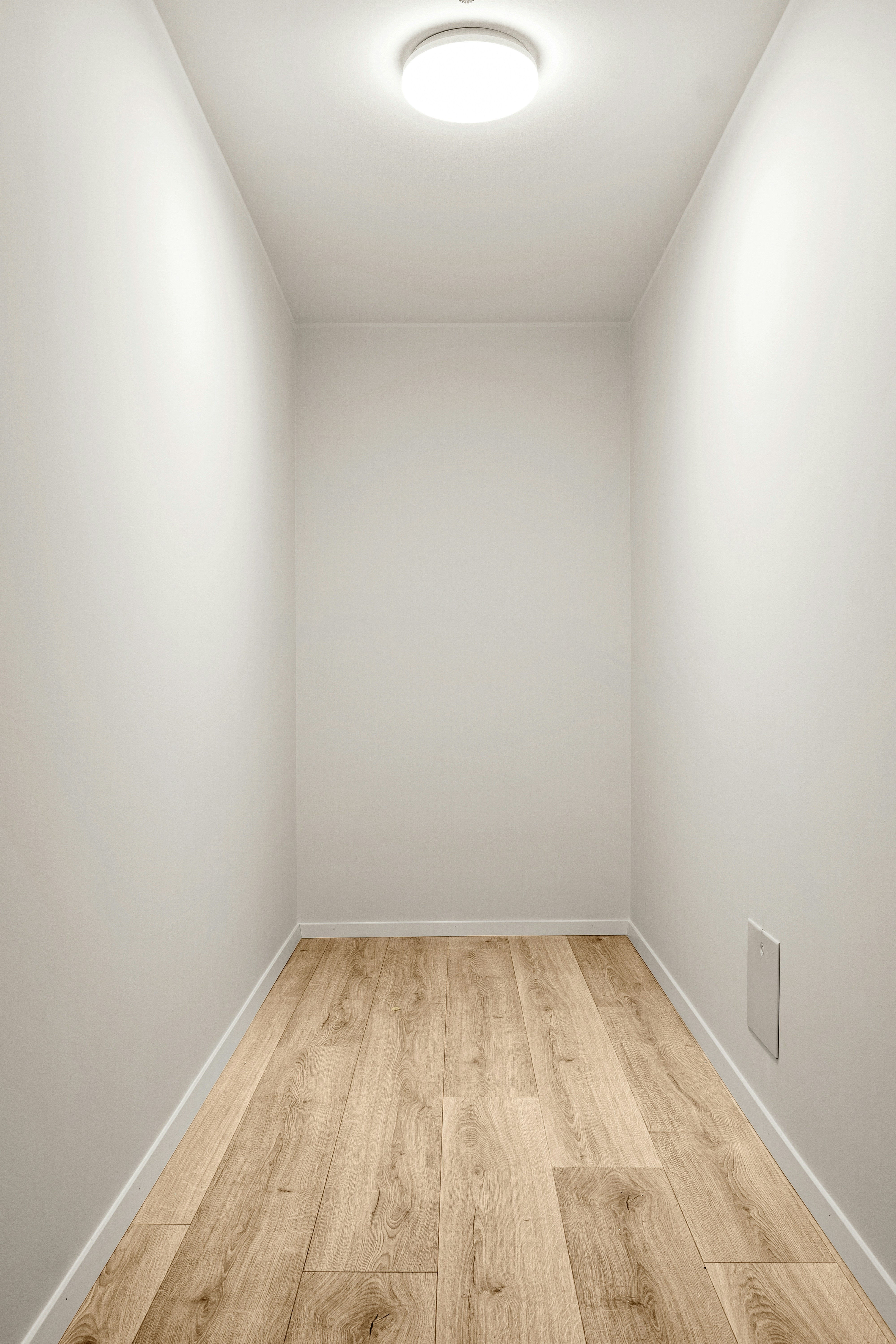 A narrow, well-lit white hallway with a round ceiling light and light wood flooring leads to a blank end wall.