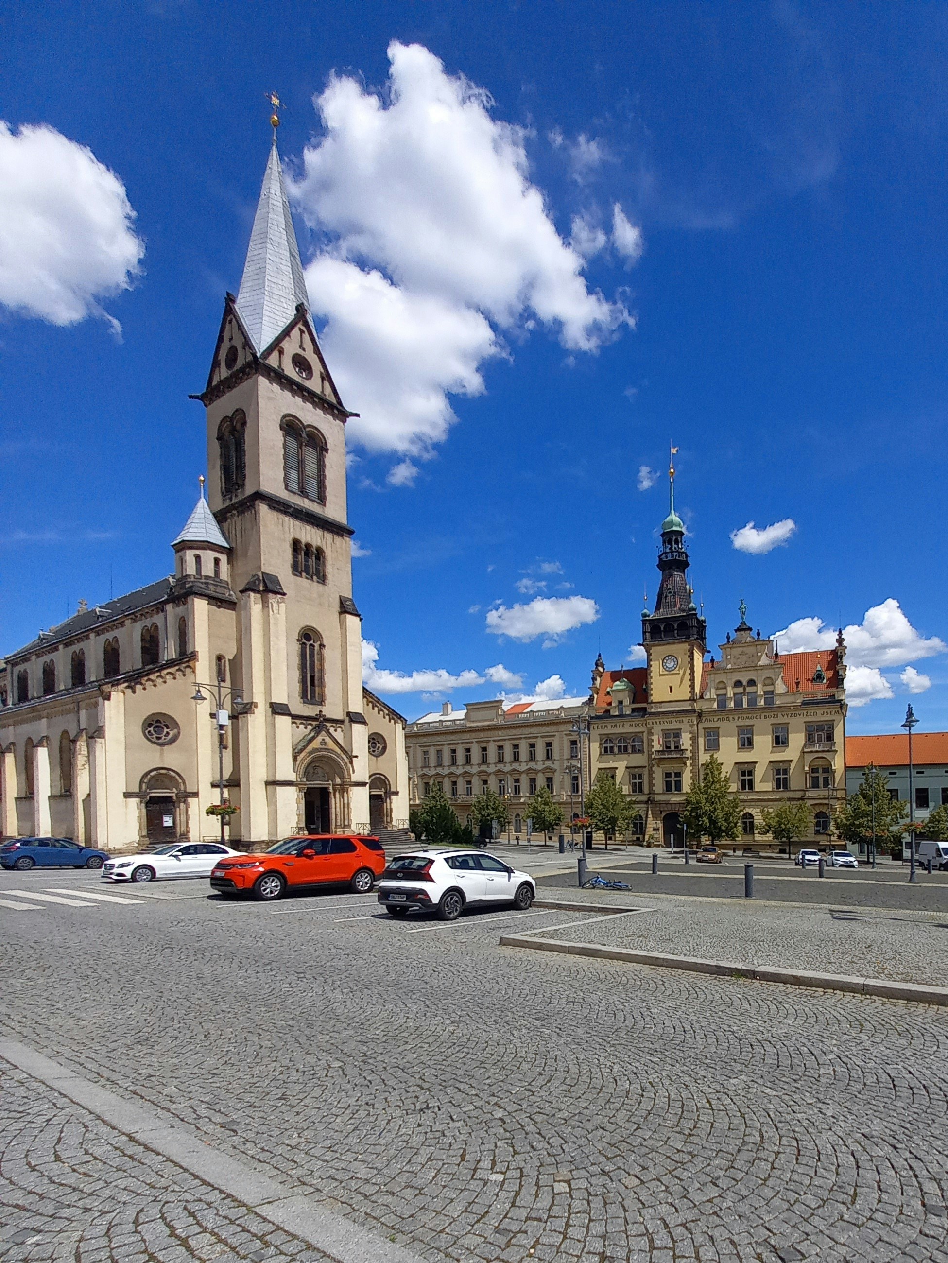 Tall church tower rises over a cobblestone square, with historic buildings and parked cars under a bright blue sky.