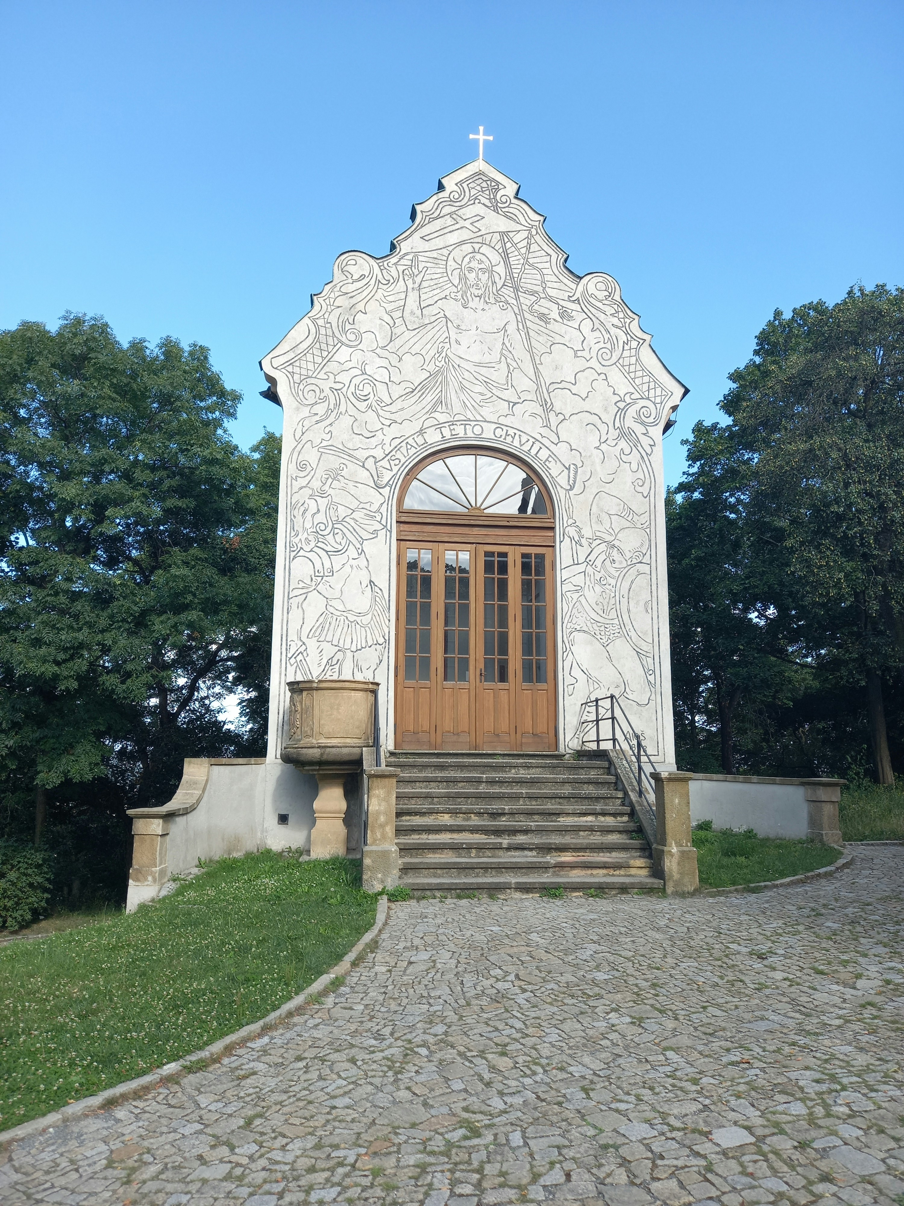 Photograph of an ornate church facade with carved reliefs and a wooden door, reached by stone steps along a cobblestone path, framed by green trees and a clear blue sky.