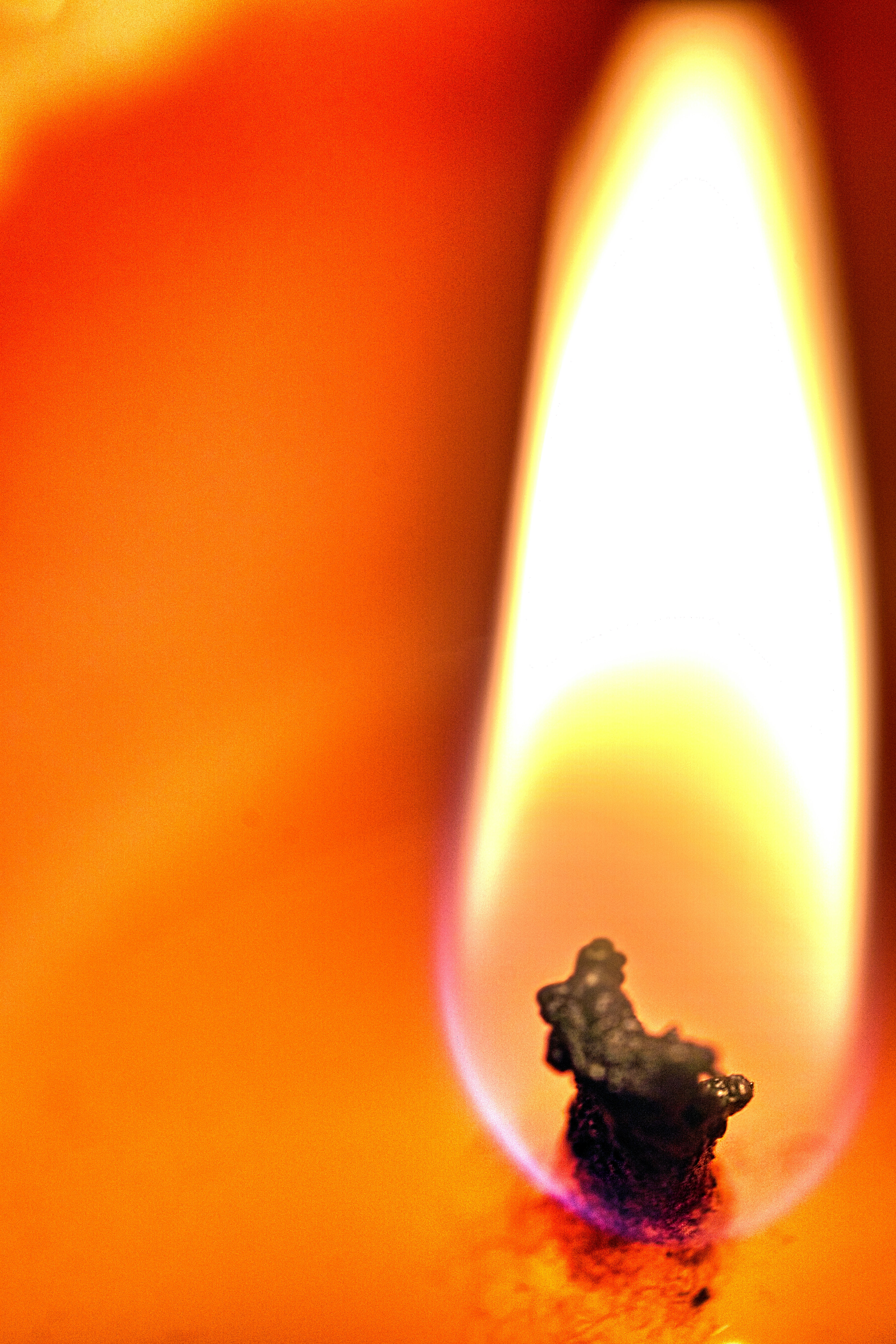 Macro photograph of a burning candle, highlighting the bright flame and charred wick against a warm orange glow.