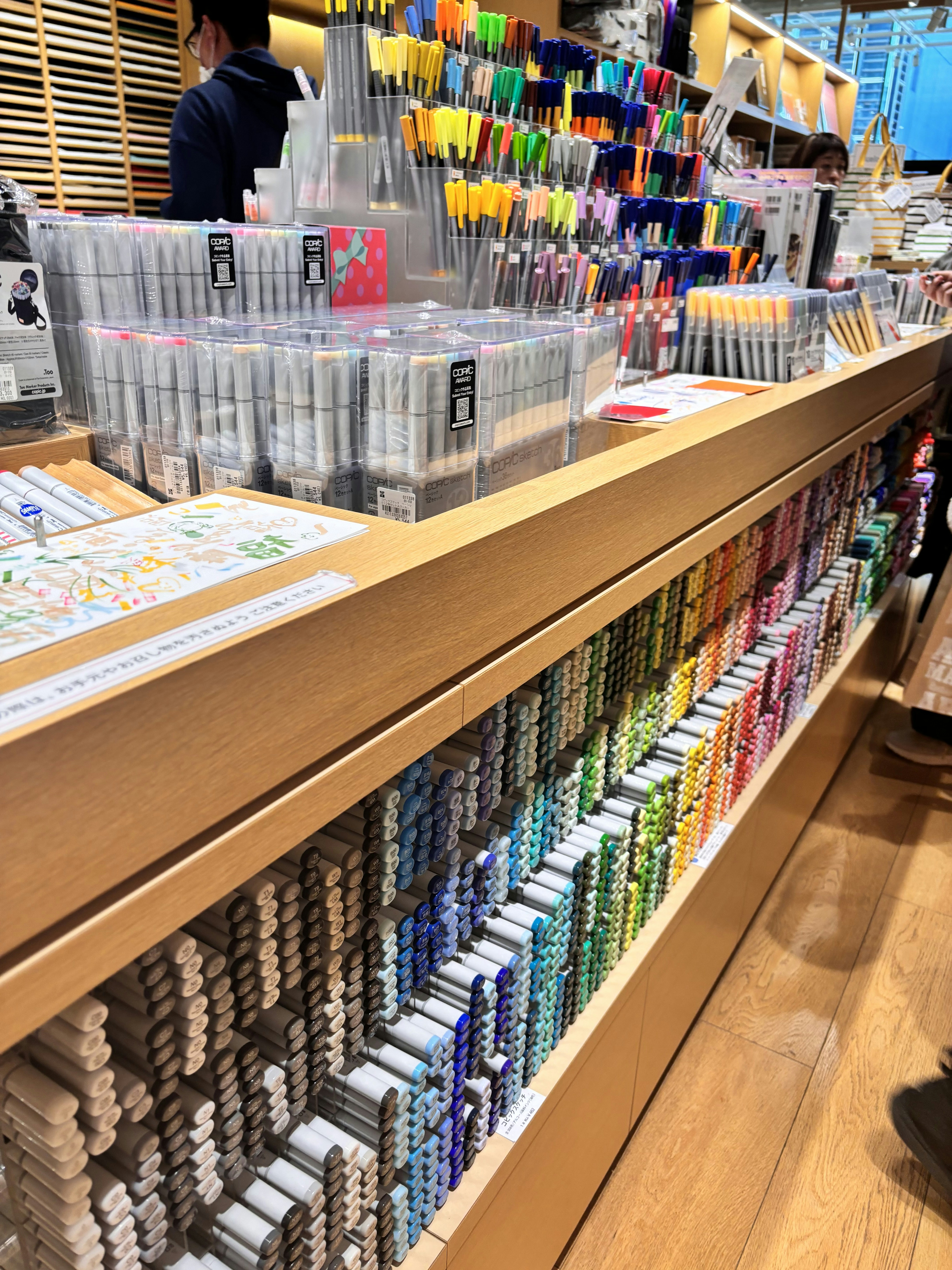Colorful marker display along a wooden counter in a stationery store, with organized rows of pens behind clear packaging.