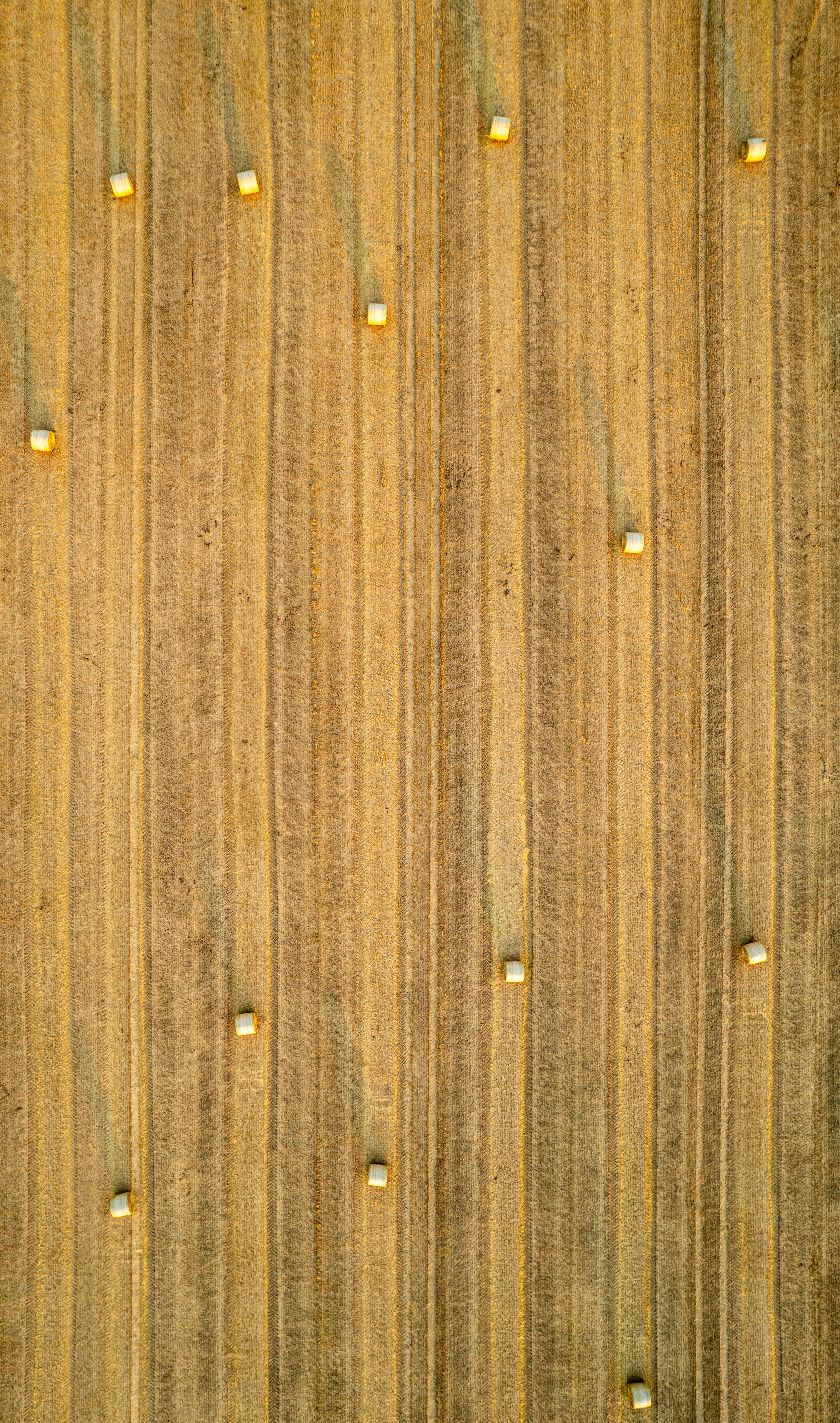 An aerial view of a harvested field featuring evenly spaced hay bales. The golden field is marked by rows of straw, creating a striking pattern. The symmetry and repetition of the hay bales and lines highlight the precision of modern agricultural practices, providing a picturesque and orderly rural landscape.