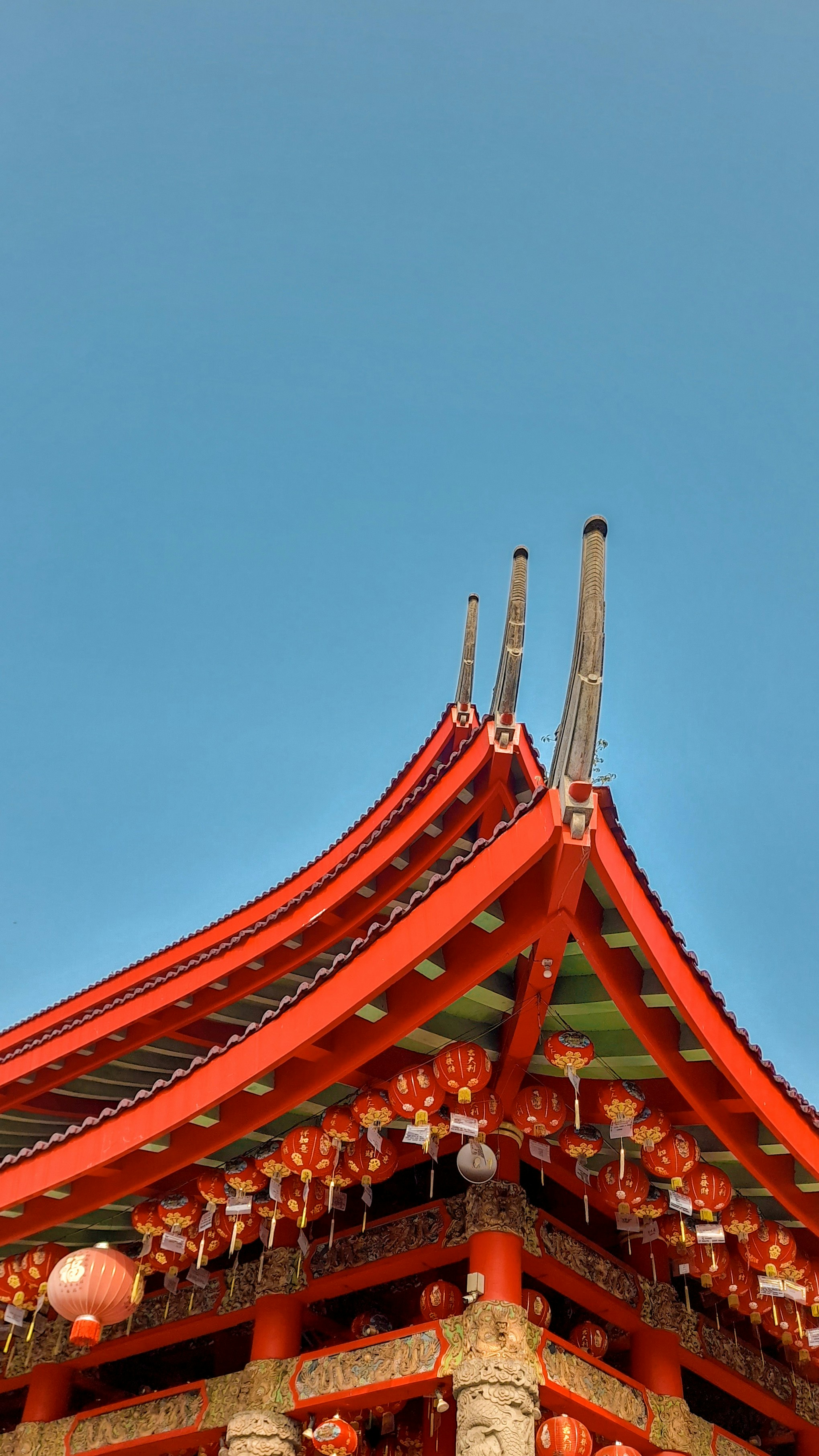 Photograph of a traditional temple roof with vivid red beams and hanging lanterns against a clear blue sky.