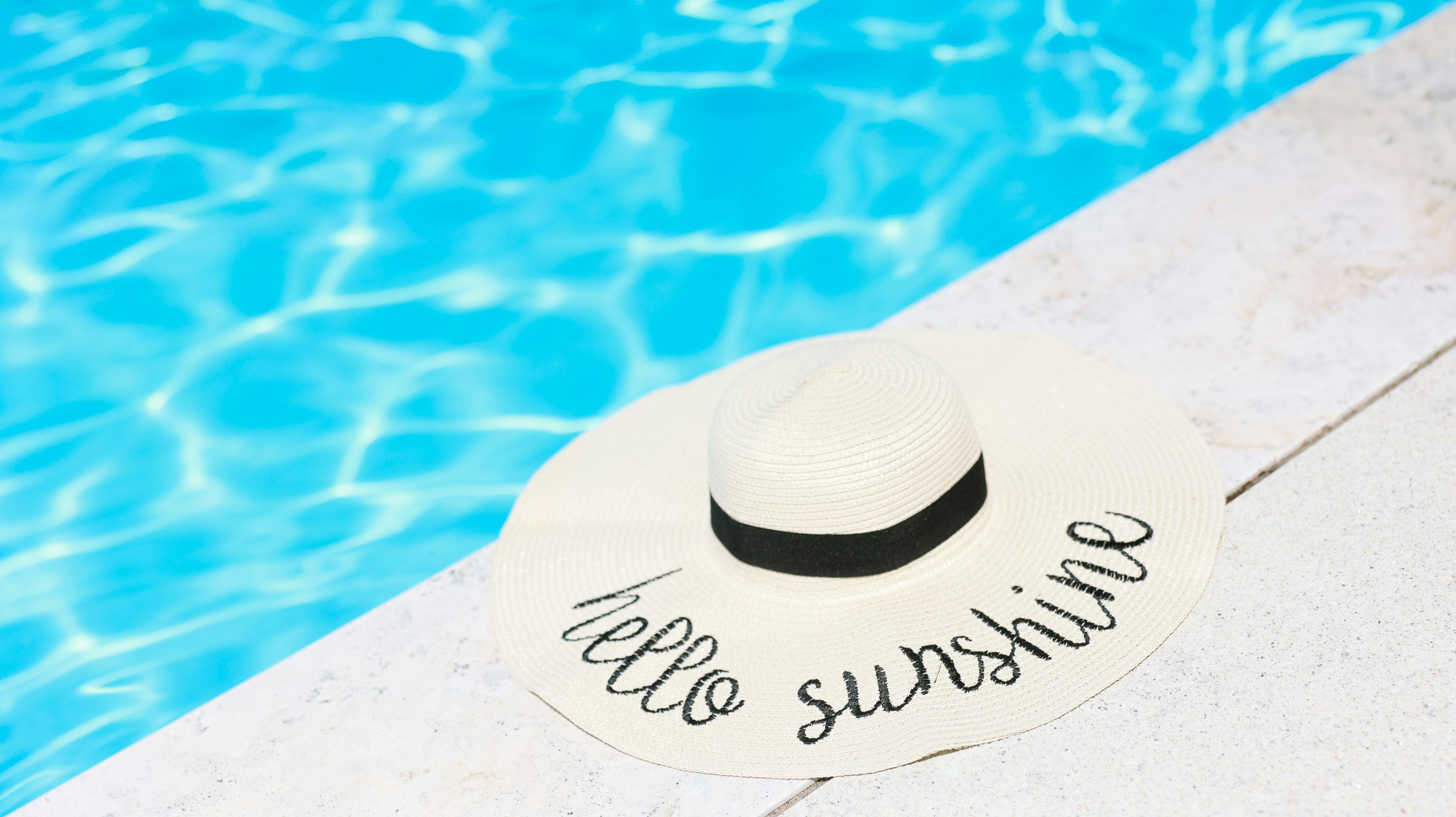 Wide-brimmed sunhat with 'hello sunshine' embroidered, resting by a shimmering blue pool. The scene evokes a relaxed summer vibe.